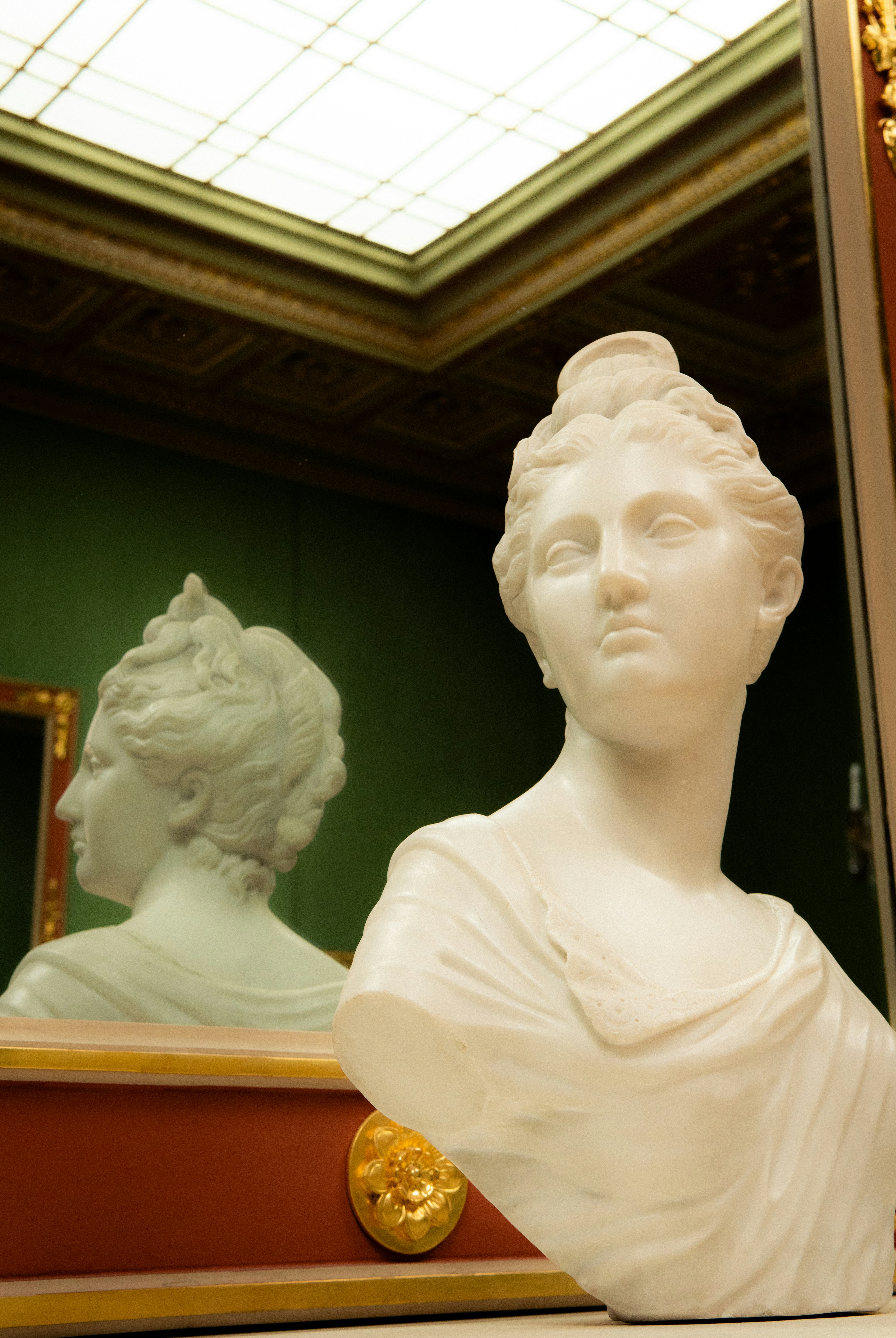 Marble bust of a classical figure, elegantly capturing the essence of beauty, with a reflection visible in a nearby mirror. The ornate surroundings enhance the historical ambiance.