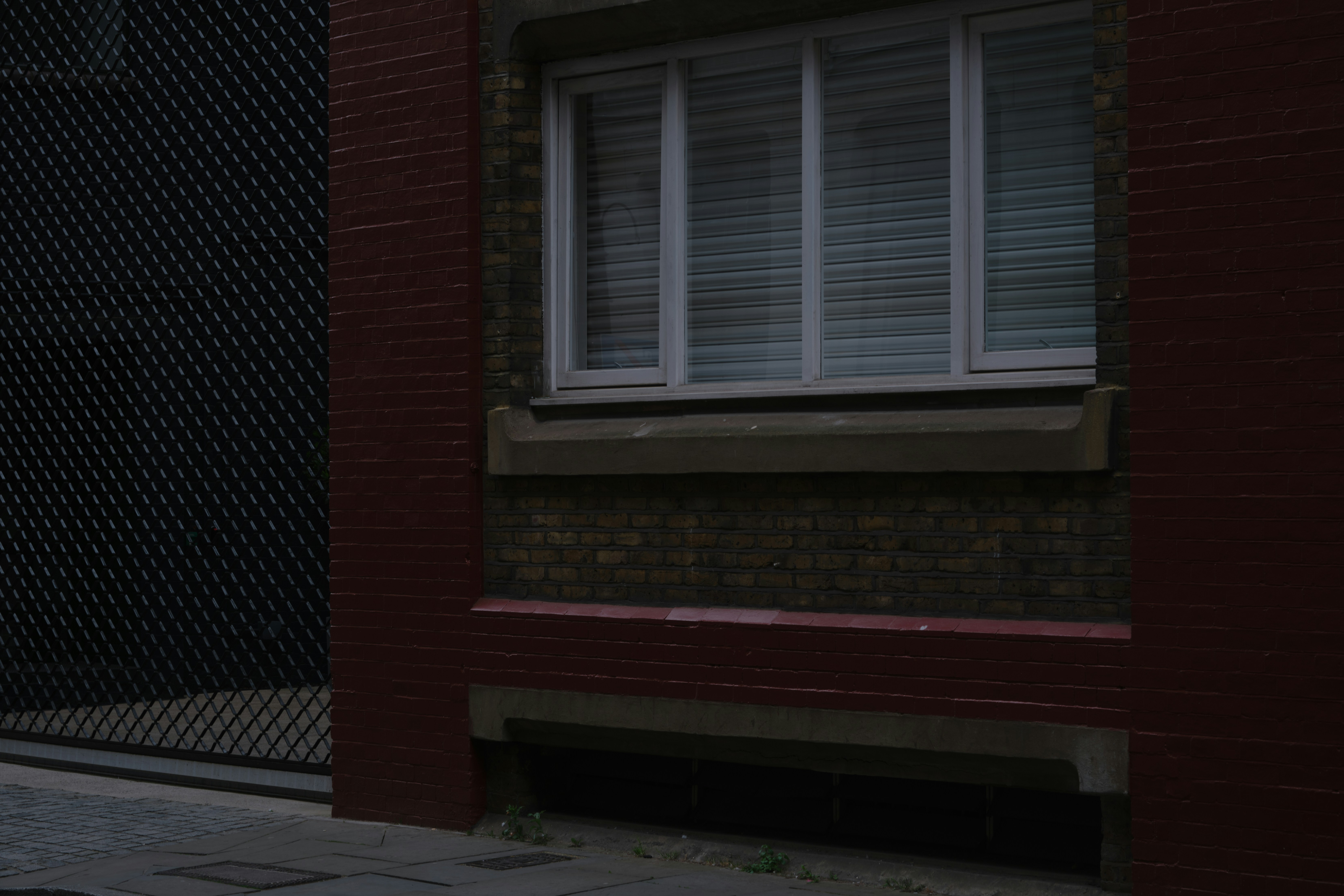A dimly lit urban scene featuring a window framed by a red wall and a black grille, hinting at the contrast between light and shadow.