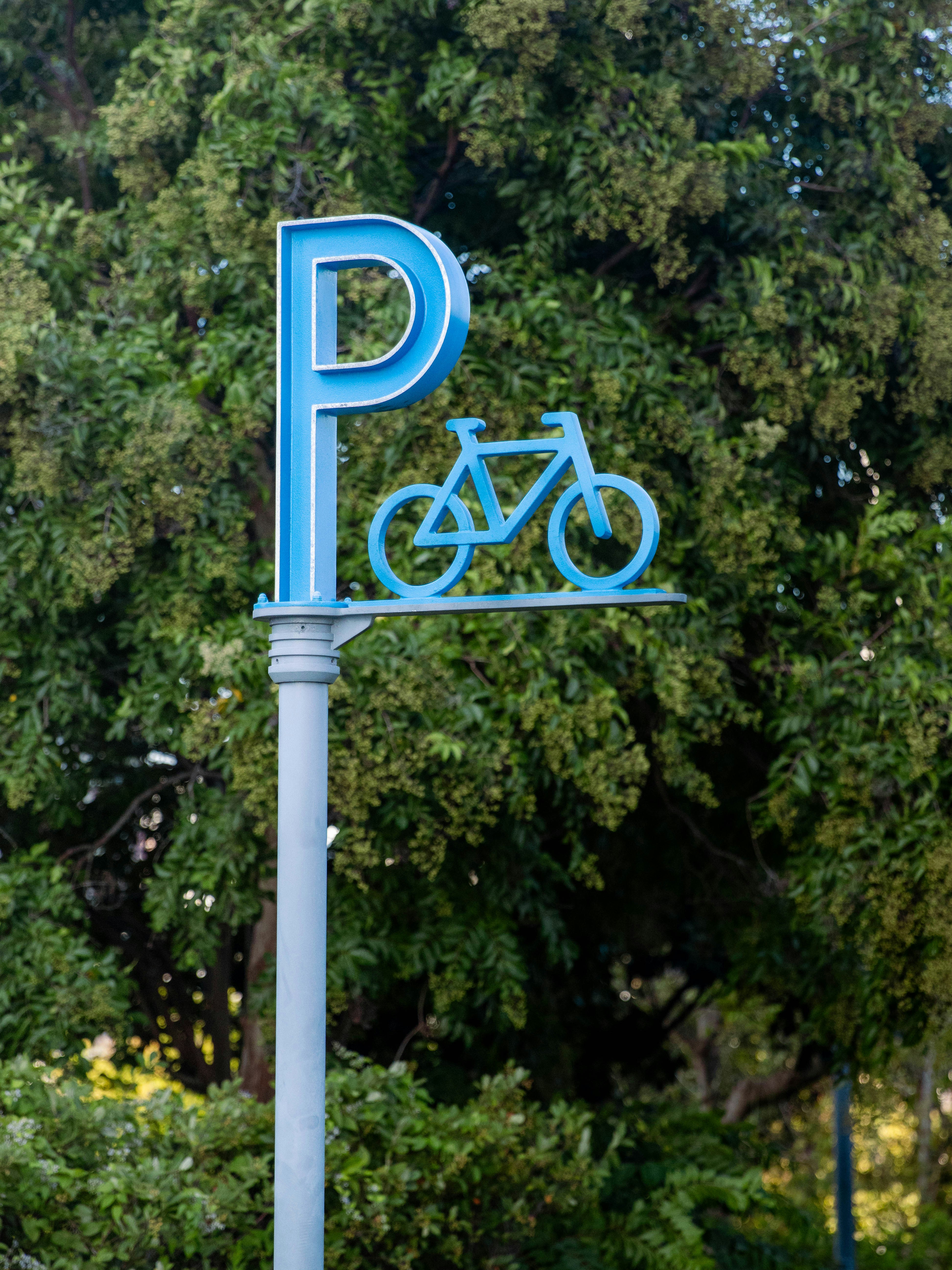 A bright blue bike and parking sign pops against leafy greenery, simple and striking. Clean setting and the color contrast make it feel friendly and easy to read. | Blue bicycle parking sign with trees in background