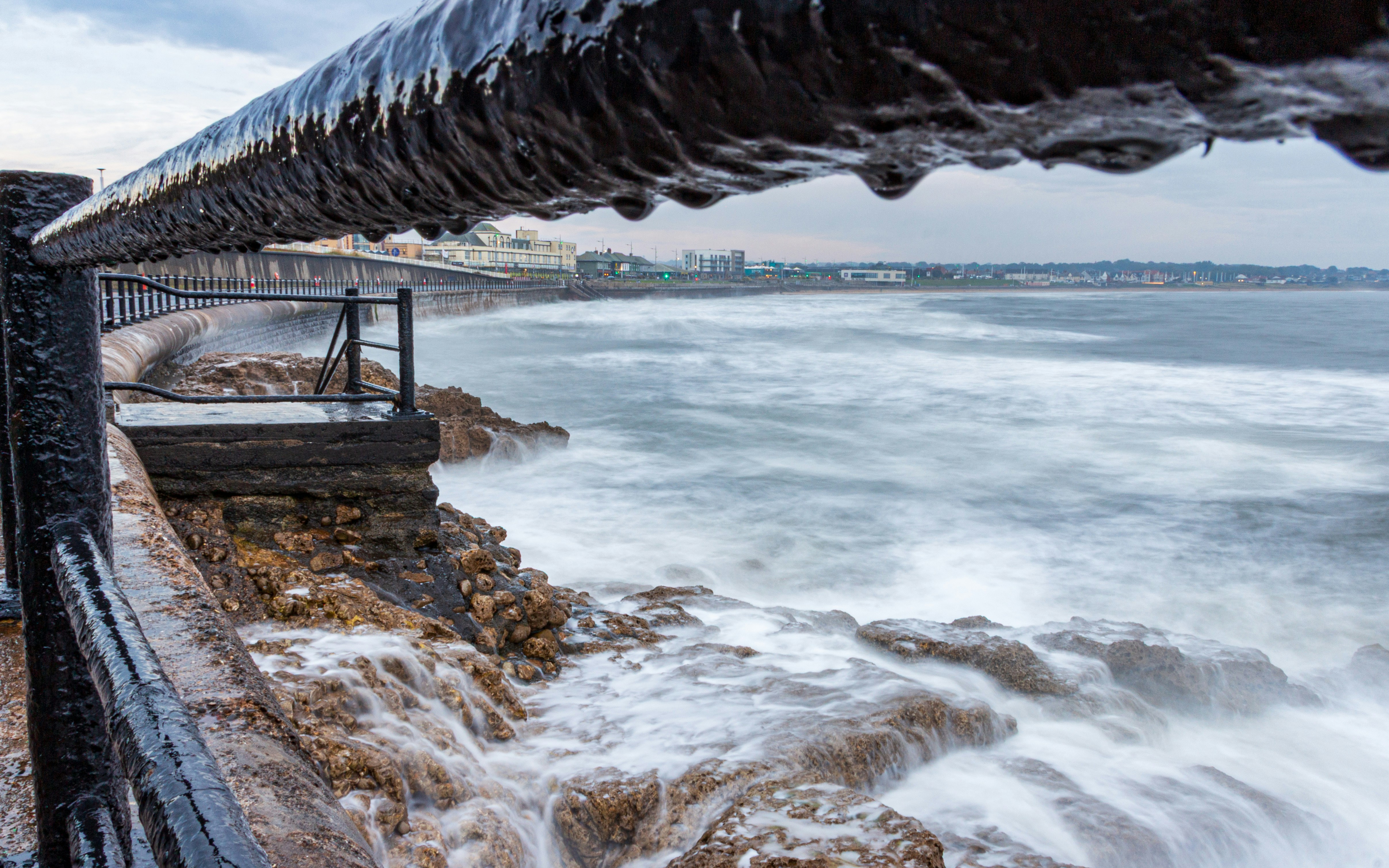 View of turbulent waves crashing against rocky shore, framed by a wet railing. The scene captures the dynamic interaction between land and sea.