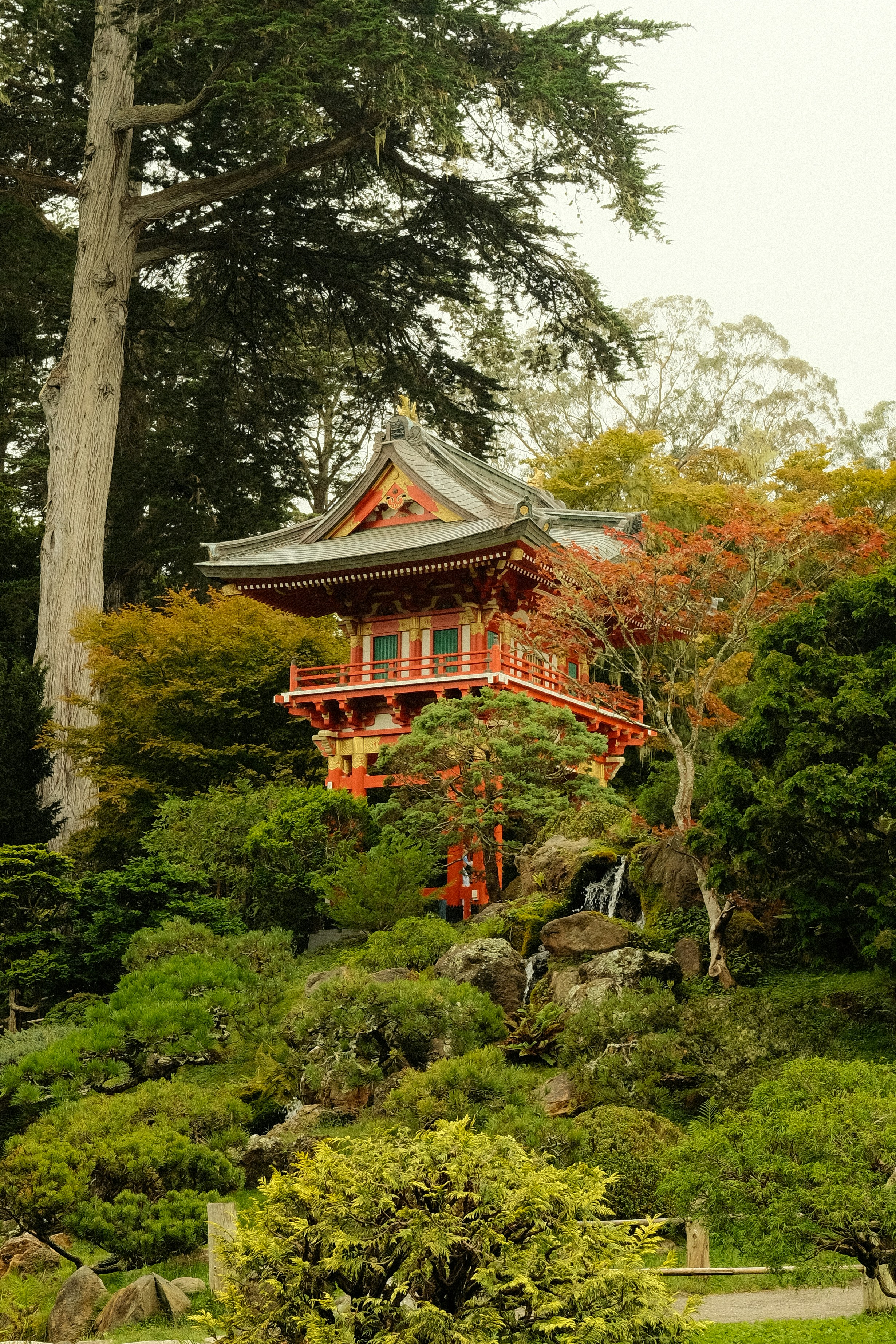 Vibrant red pagoda nestled among lush greenery and gentle waterfalls in a tranquil garden setting.