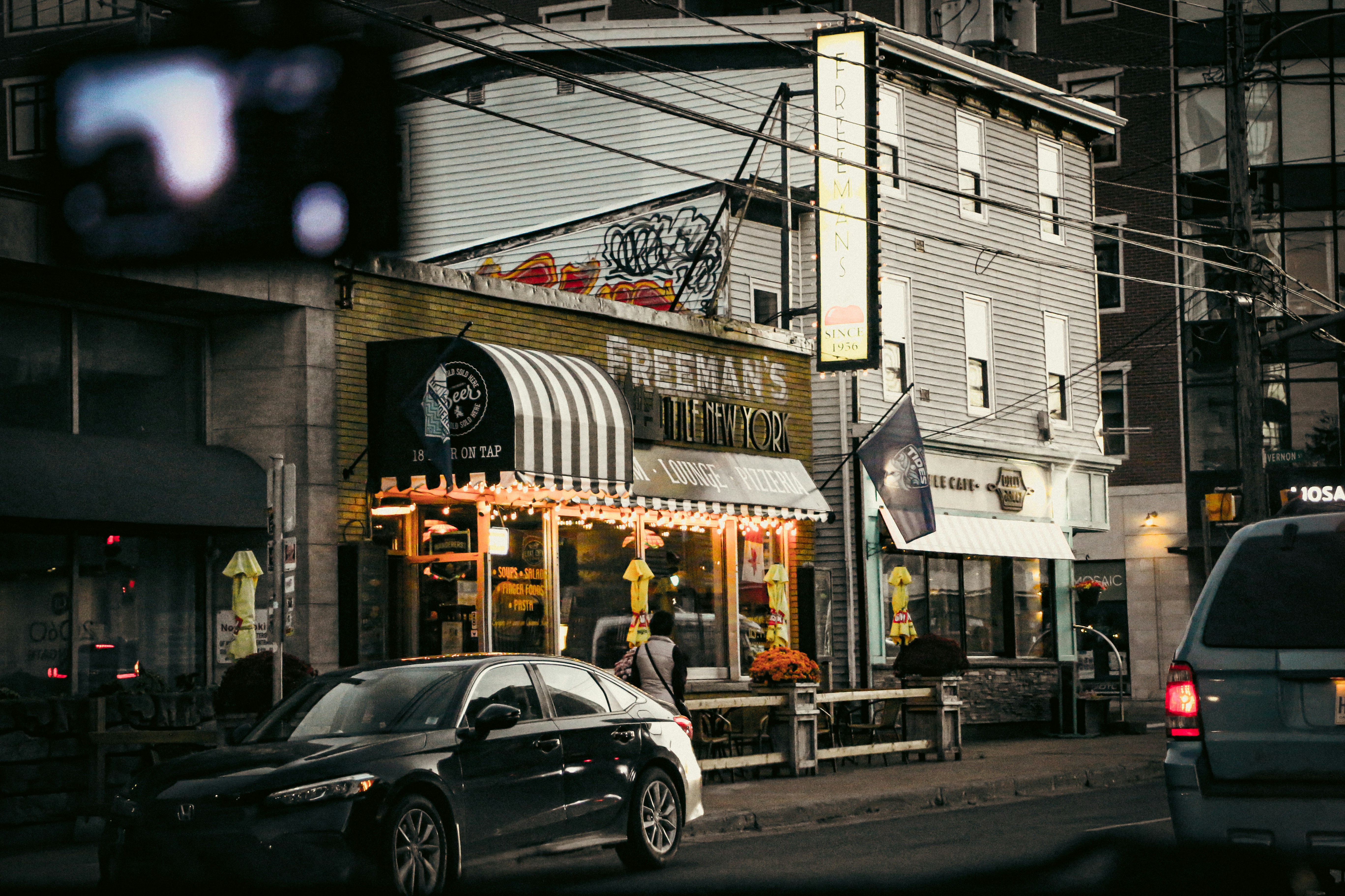 Freeman's Eatery glows warmly under evening lights, inviting patrons with its vibrant signage and outdoor seating. A car passes by, adding to the urban ambiance.