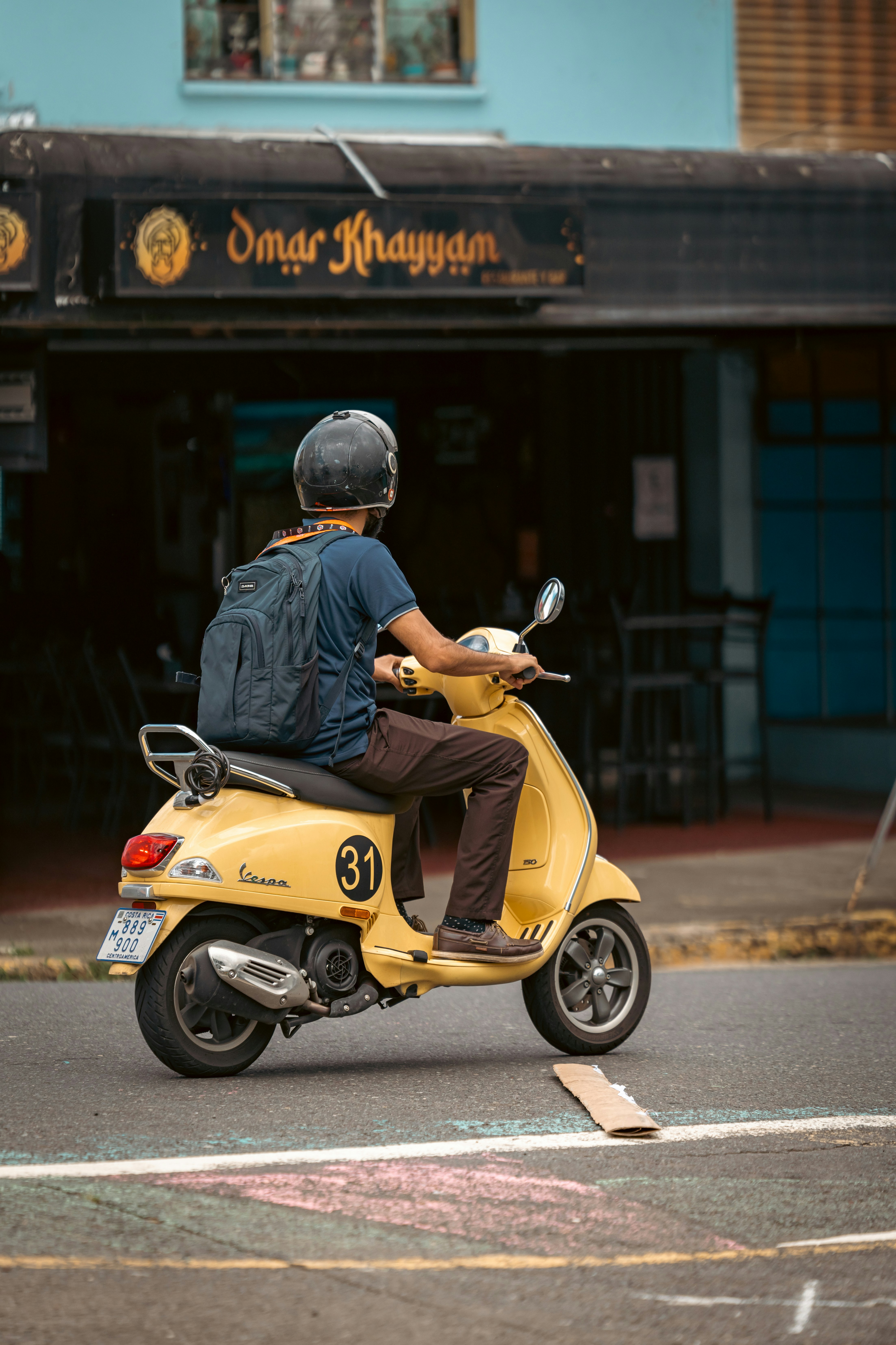 Par de una sesión de street photography en San Pedro, San José, Costa Rica | Man riding a yellow vespa scooter past a building.