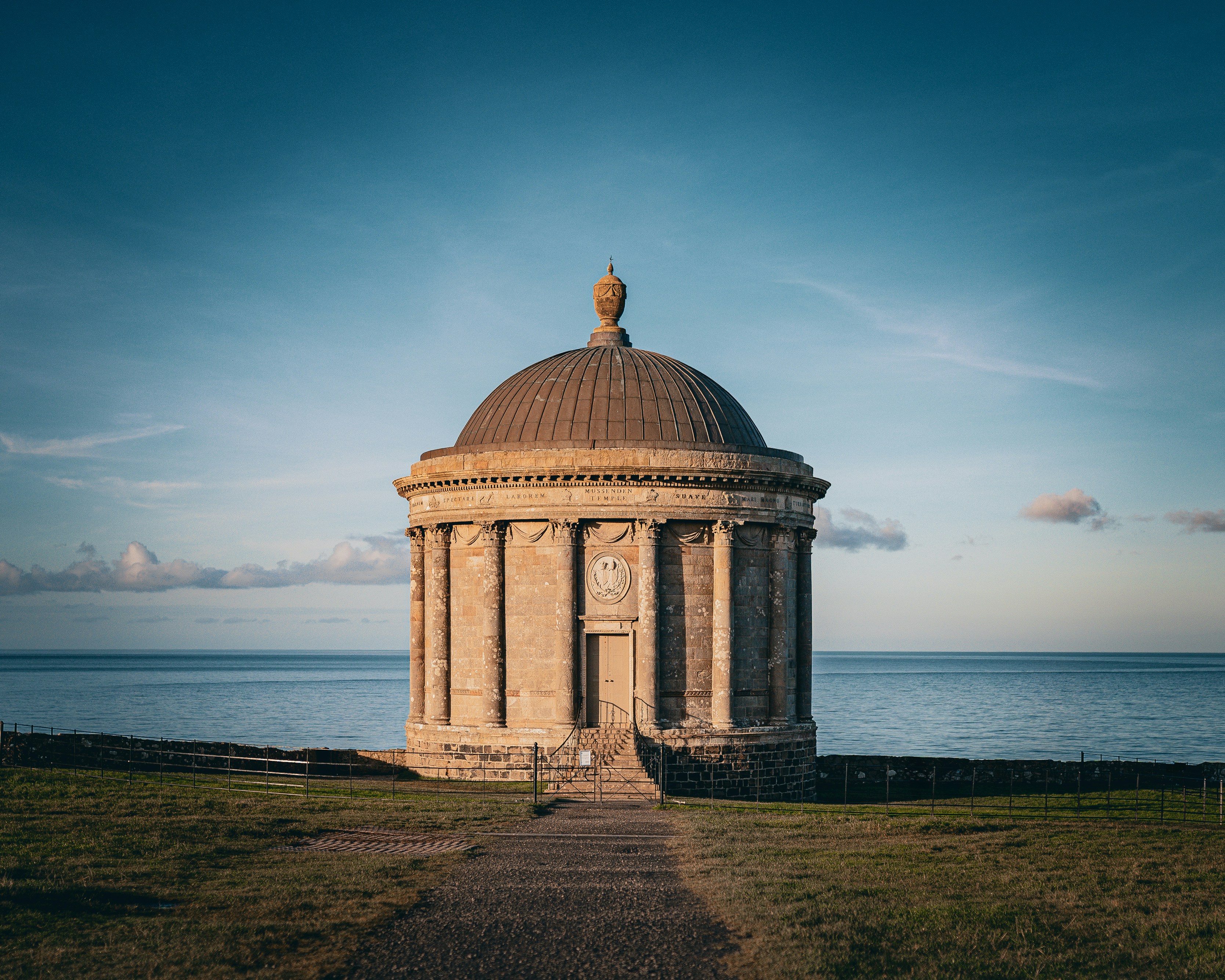 A historic pavilion stands majestically by the shoreline, framed by a tranquil sea and a clear sky. The structure's architectural details are highlighted in the soft evening light.