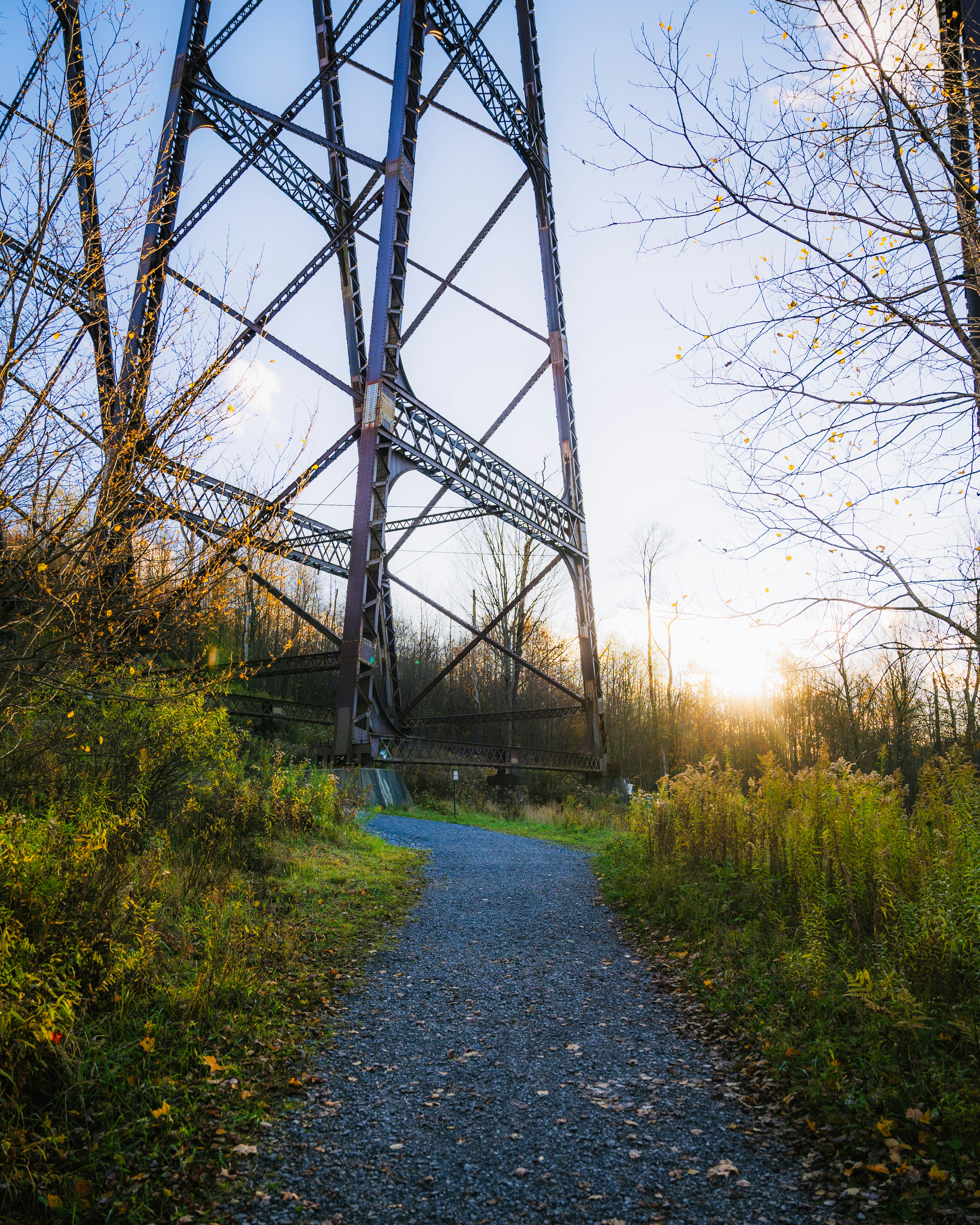 A gravel pathway leading towards a towering metal structure amidst a tranquil landscape with autumn foliage. The setting sun casts a warm glow over the scene.