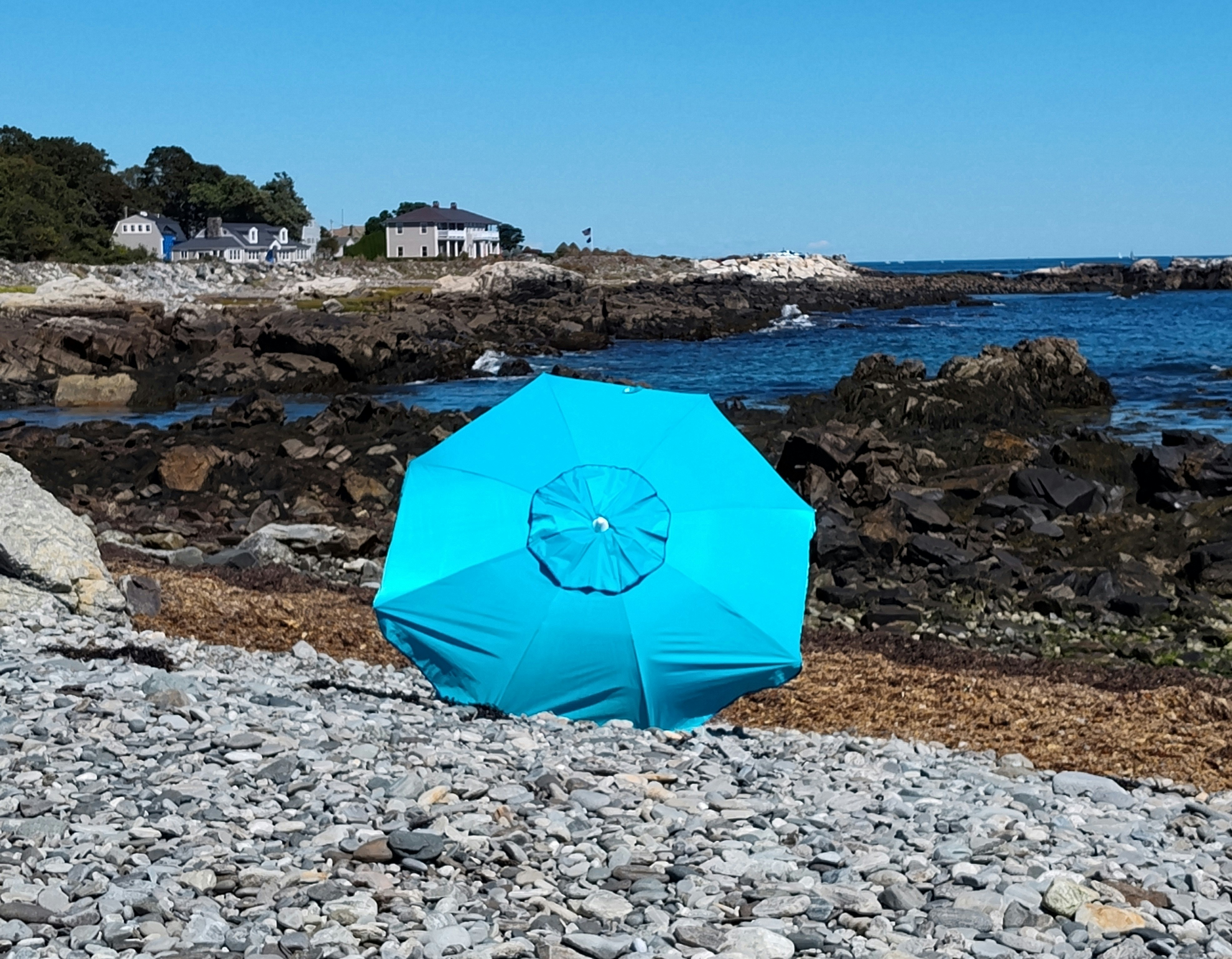 Bright turquoise umbrella set against a rocky beach with coastal homes in the background. The scene captures a tranquil day by the water.