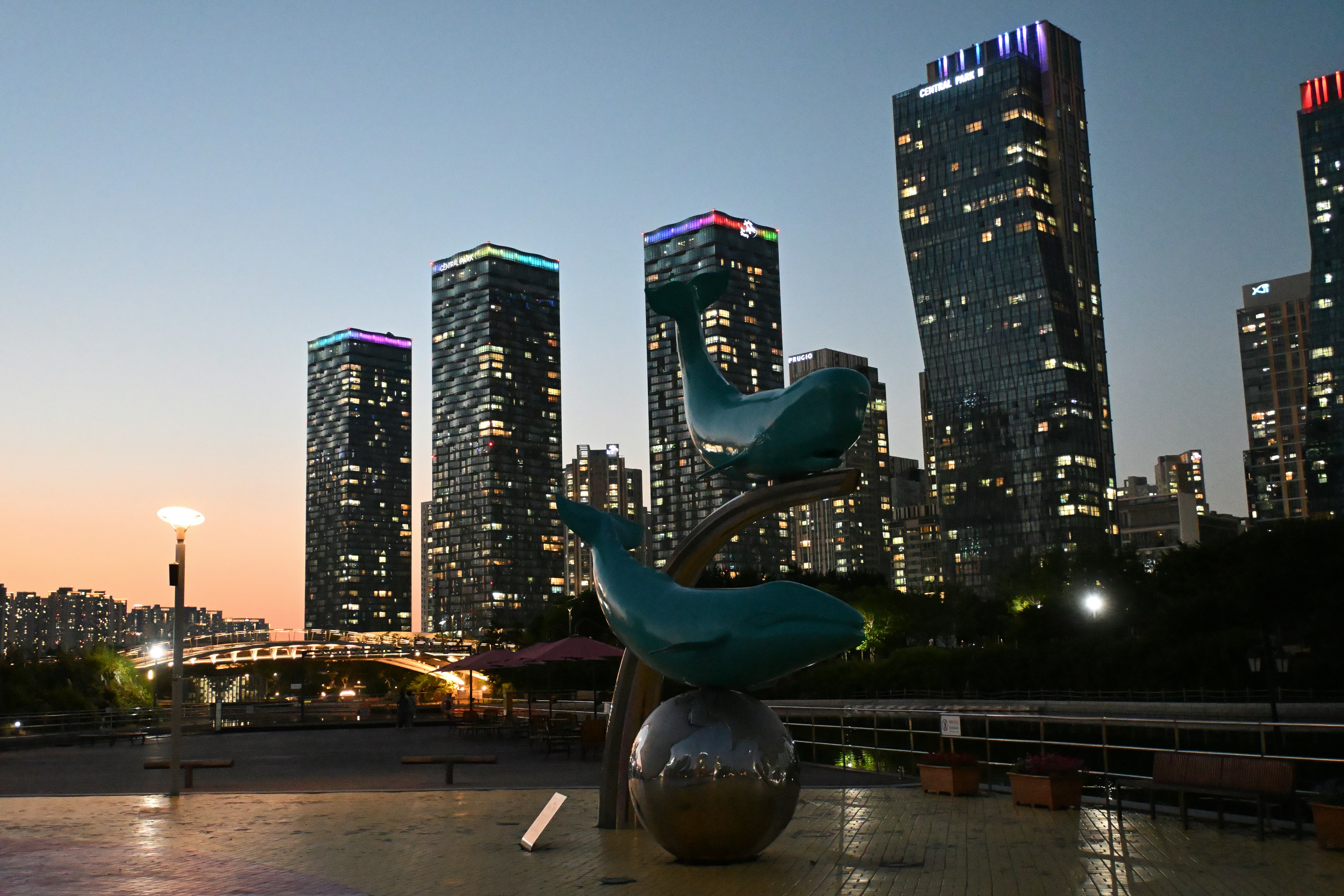 Teal Bird Sculptures Stand Guard Over a Dusk-Glowing Riverside Skyline