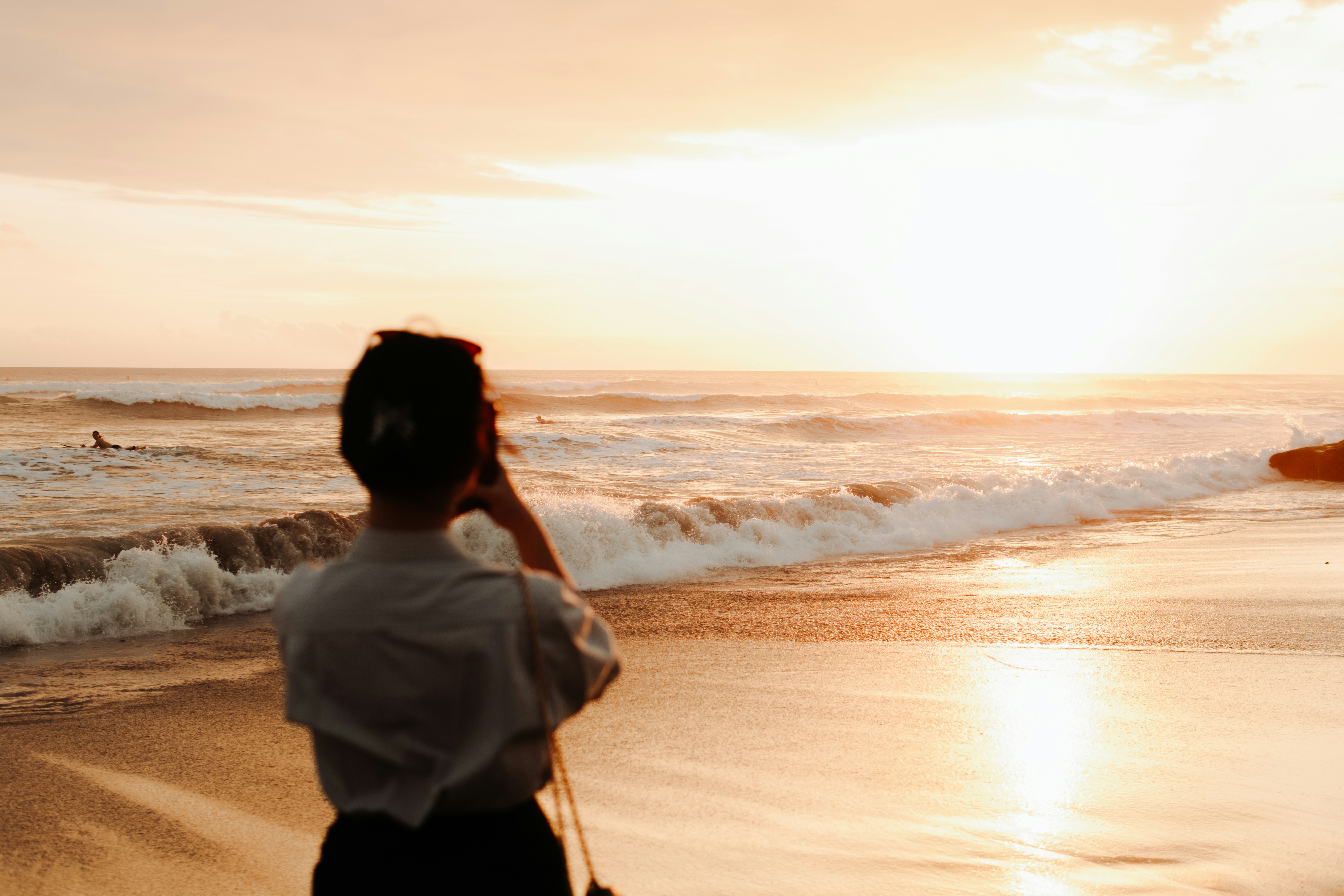 Silhouetted figure capturing the sunset at a tranquil beach, waves gently lapping at the shore. The warm glow of the sun reflects off the water.