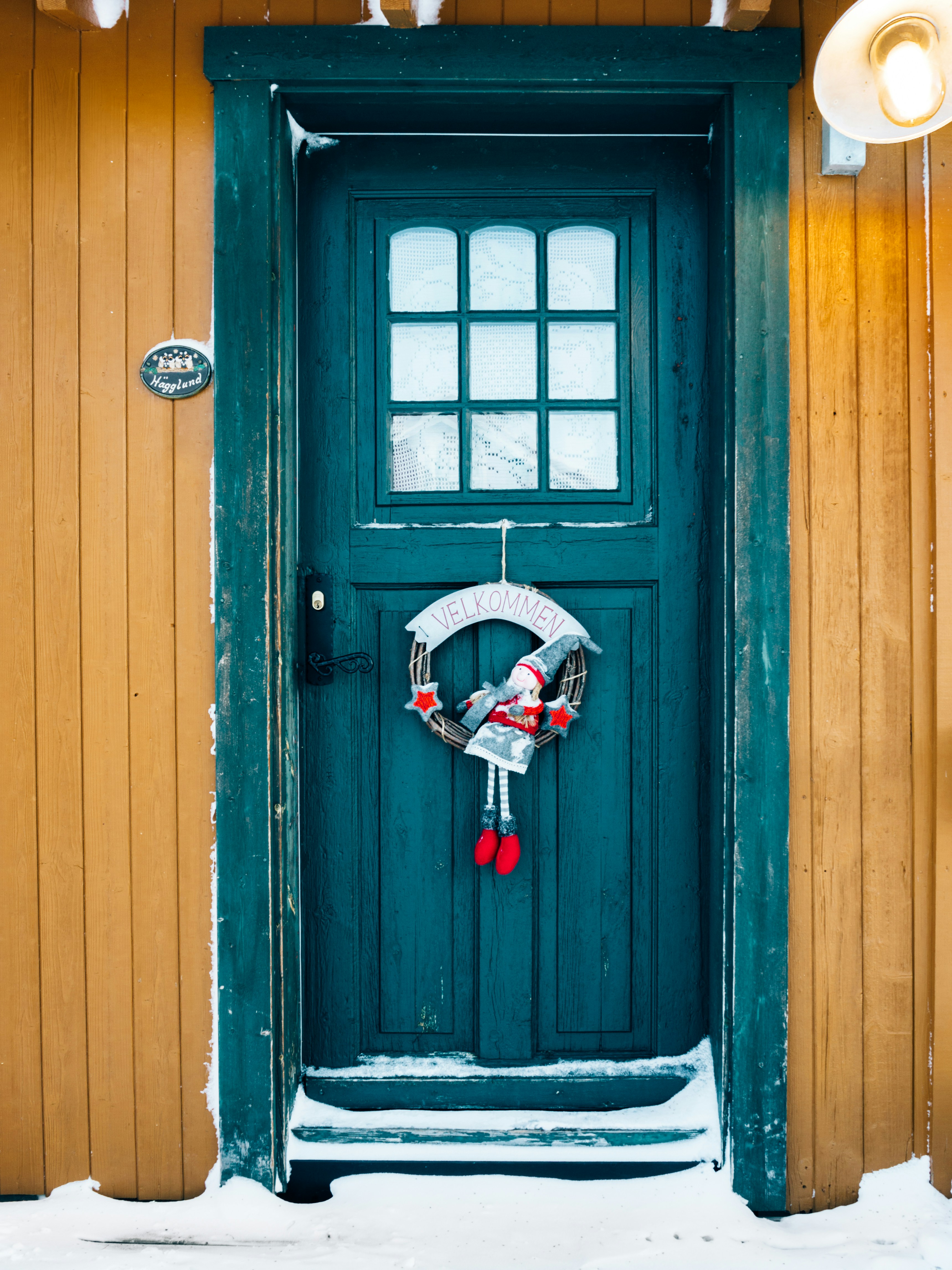 A charming teal door adorned with a festive wreath and a playful decoration, set against a rustic wooden wall. Snow blankets the ground, enhancing the cozy atmosphere.