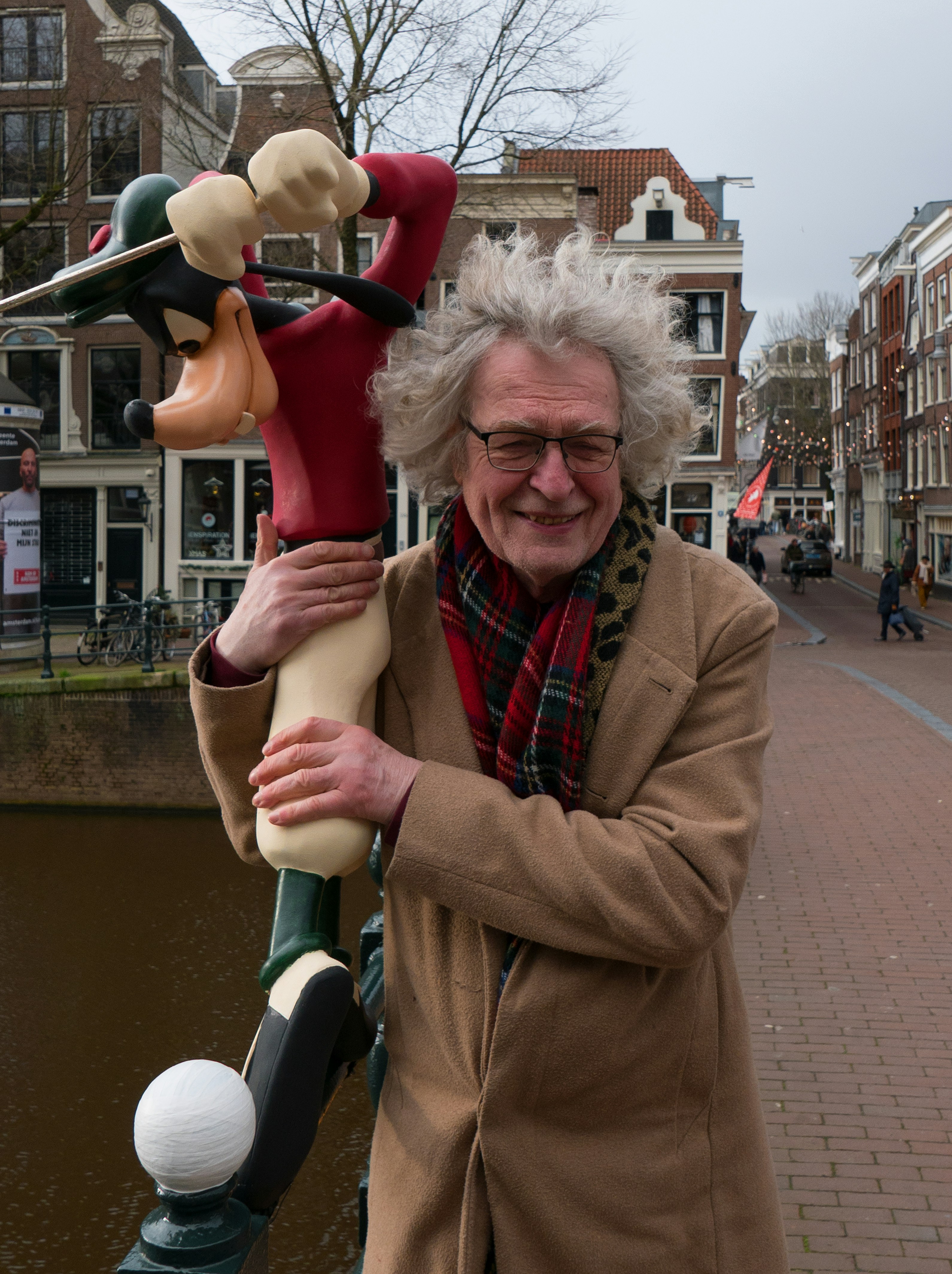 An elderly man joyfully poses with a Goofy statue along a canal, showcasing a blend of humor and nostalgia. The urban backdrop adds to the lively atmosphere.