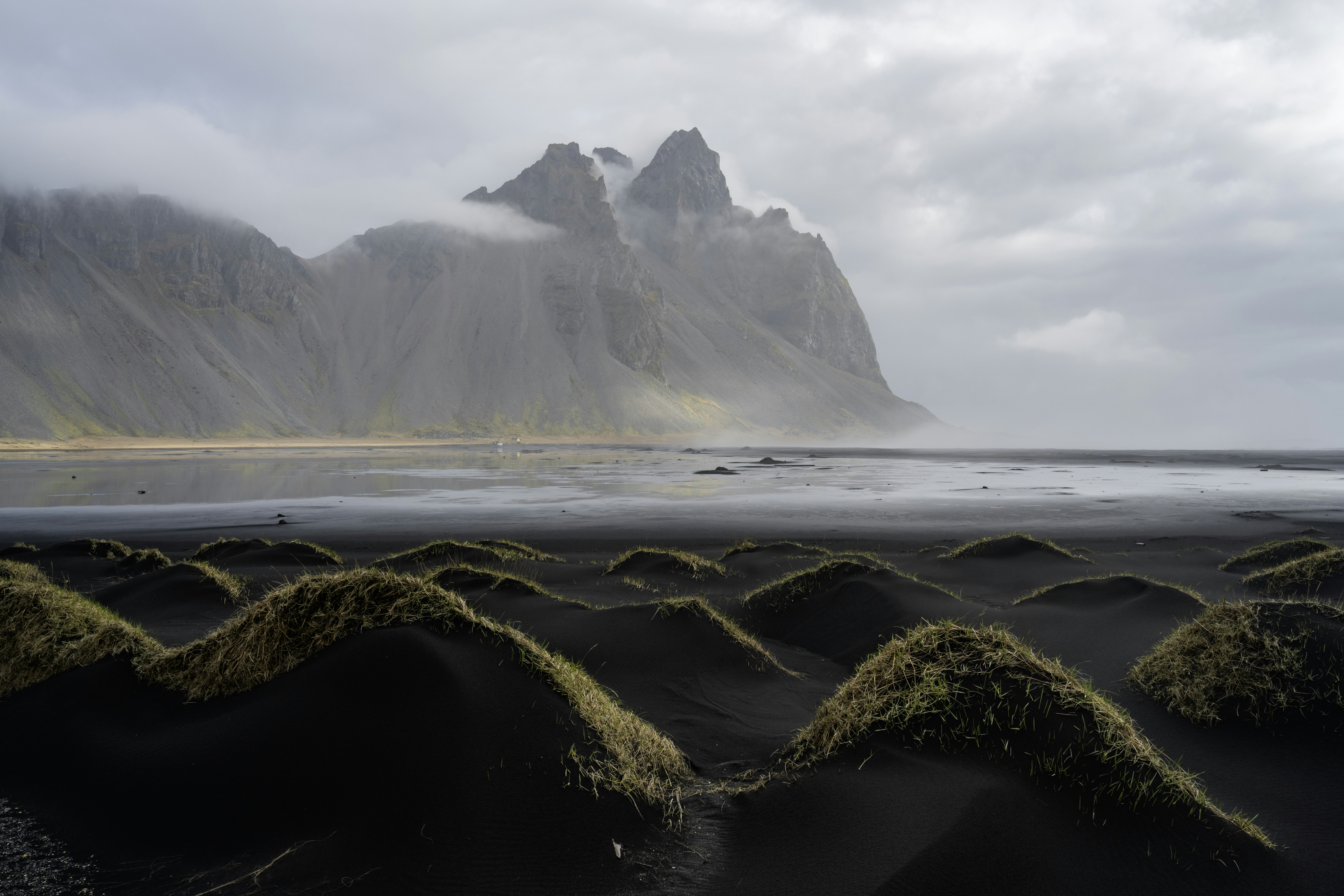 Mysterious black sand dunes adorned with tufts of grass, set against a backdrop of towering, mist-covered mountains. 