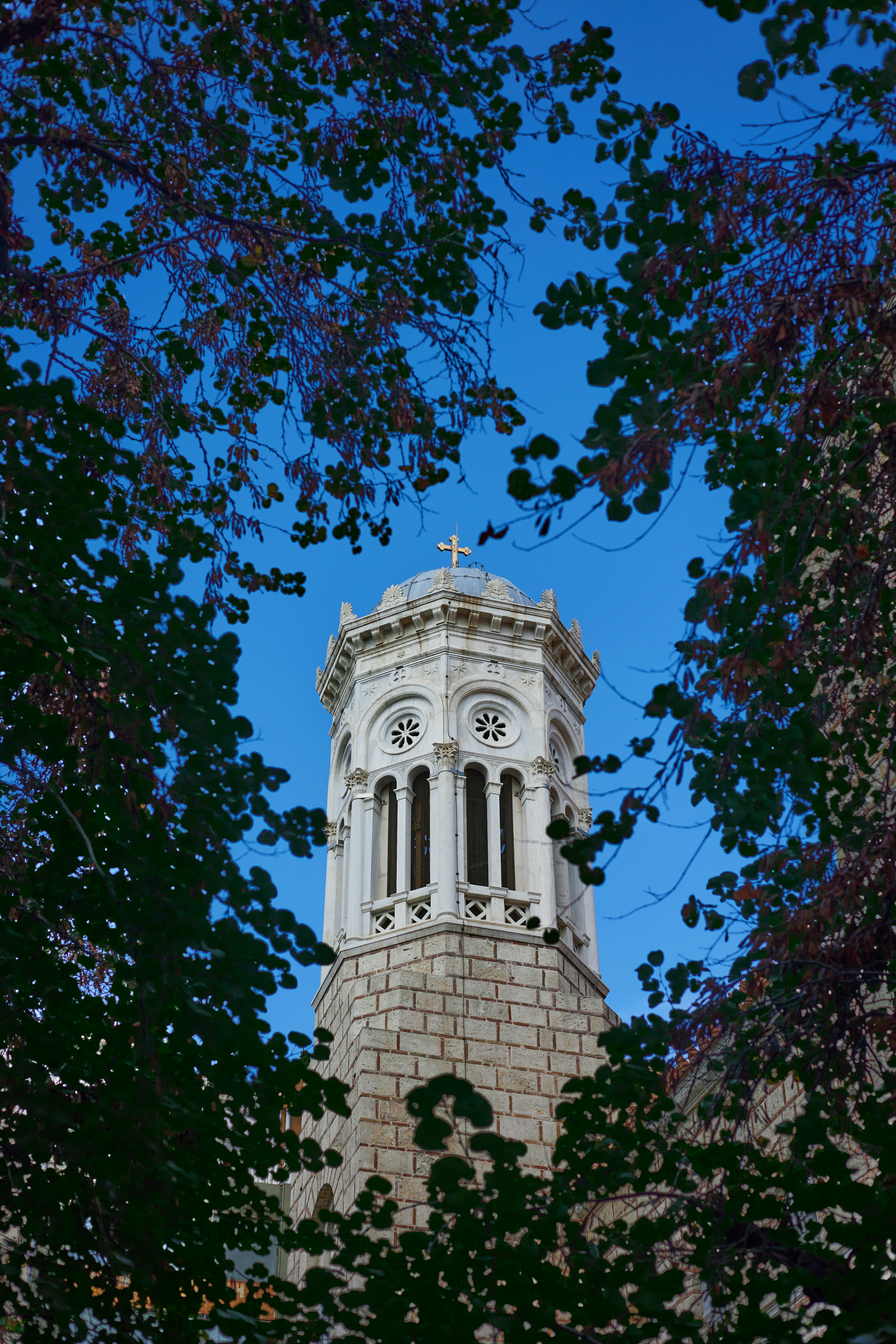 A church tower rises majestically against a clear blue sky, framed by lush green foliage. The intricate architectural details of the tower are highlighted in the sunlight.
