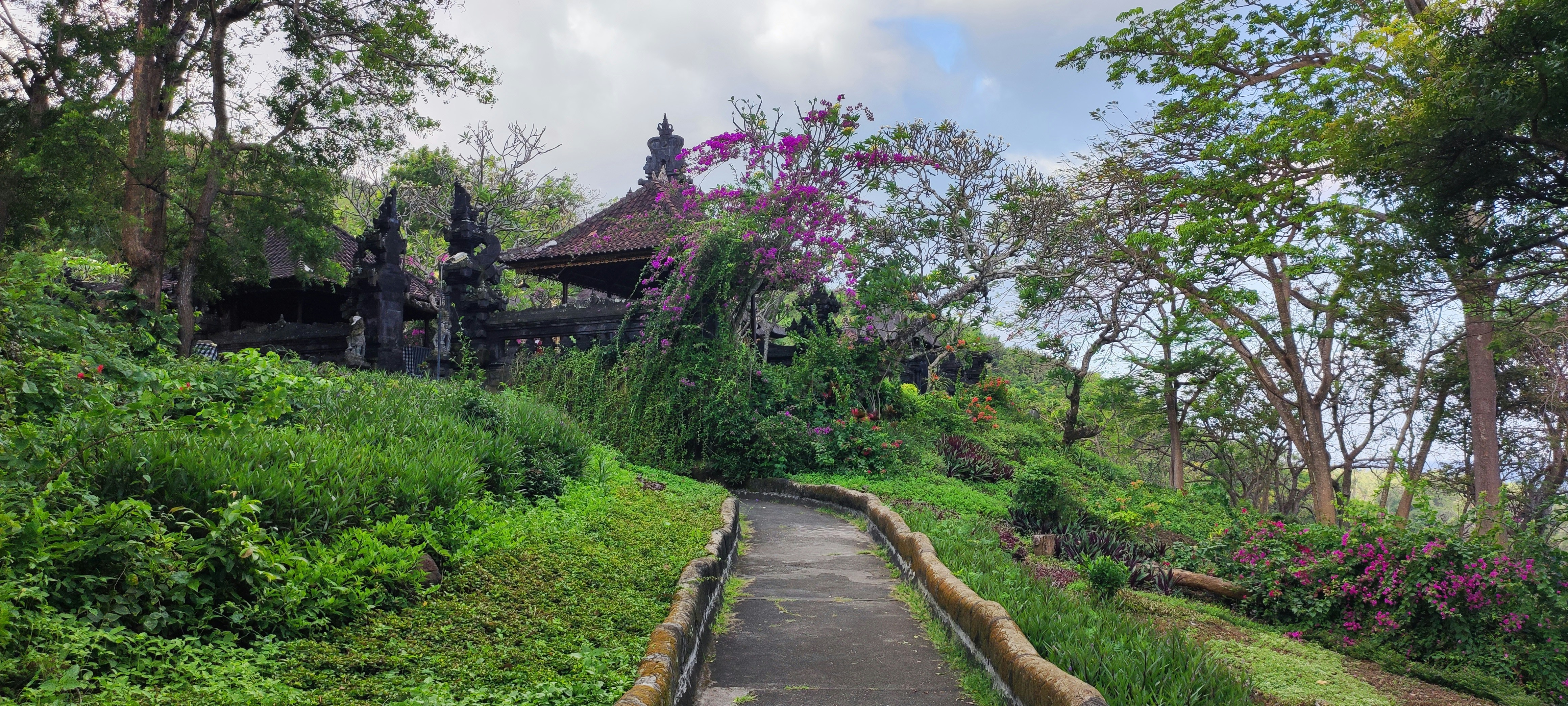 Balinese temple surrounded by tropical vegetation and colorful flowers, scenic pathway leading uphill to sacred site, traditional architecture in lush nature, cultural landmark in Bali, Indonesia. | Pathway leading to a tropical temple with lush greenery.