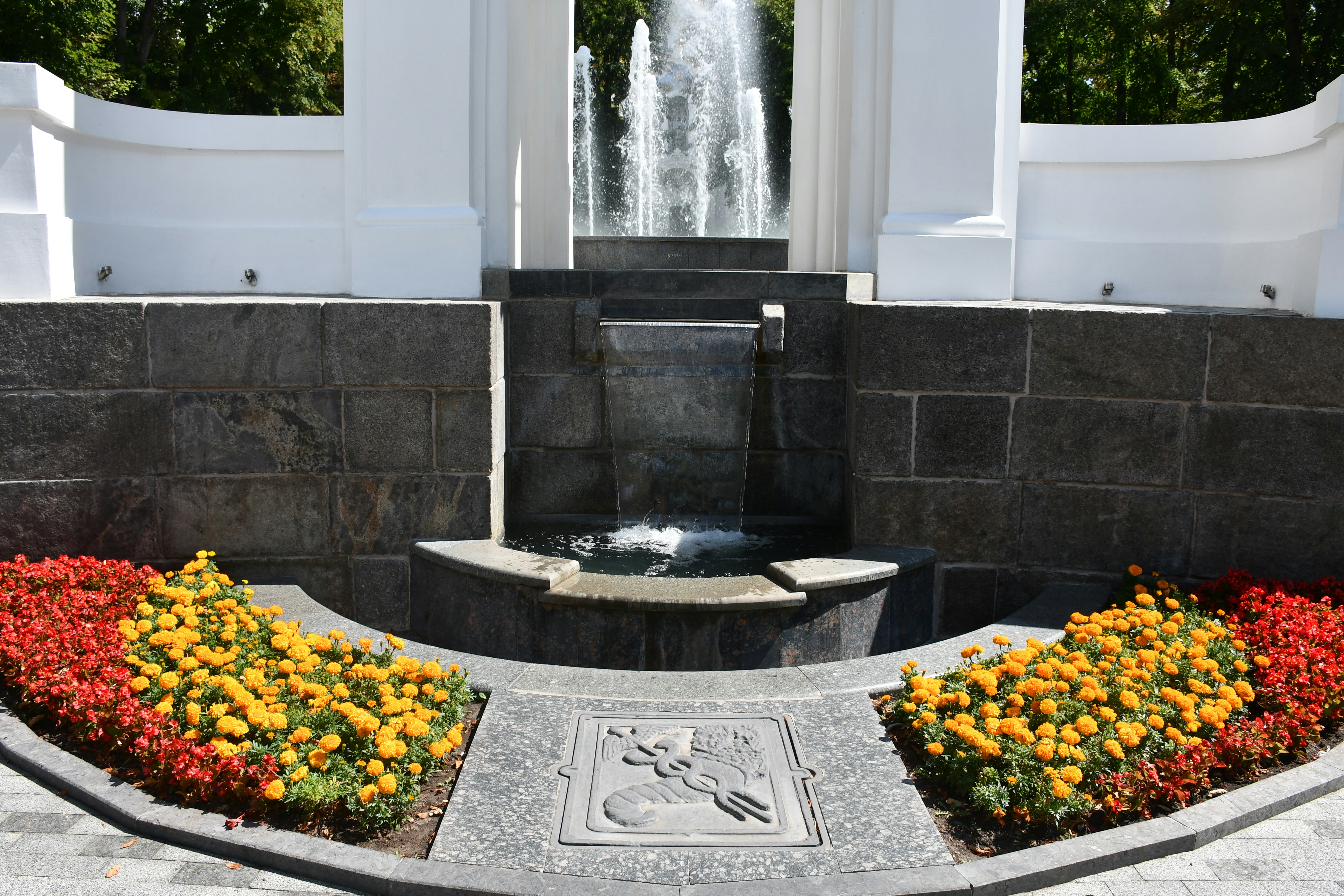 Symmetric White Arch Fountain Framed by a Stone Wall and Vibrant Red and Yellow Flowerbeds