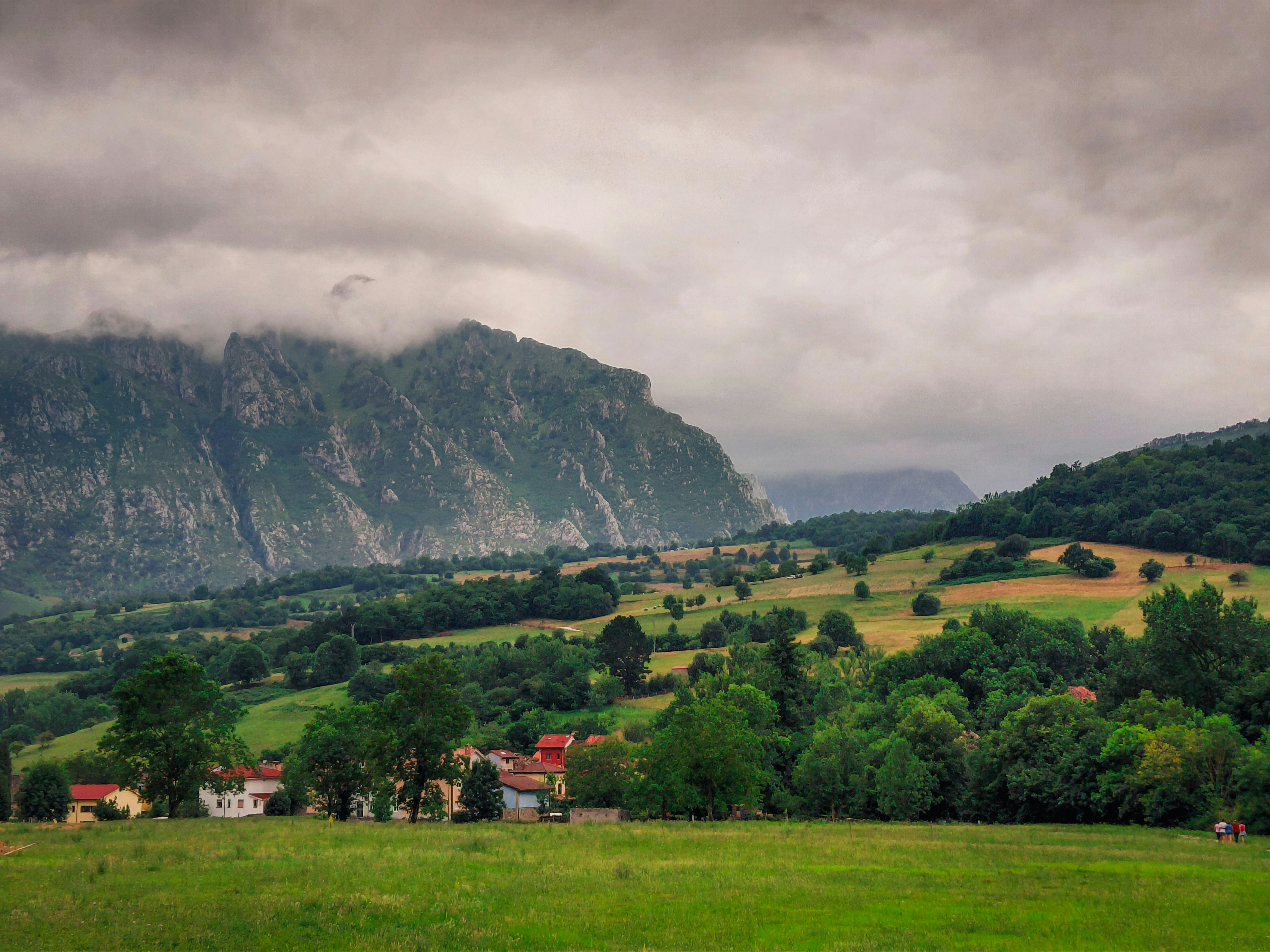 Lush green fields stretch towards majestic mountains shrouded in clouds, revealing a quaint village nestled in the valley. 