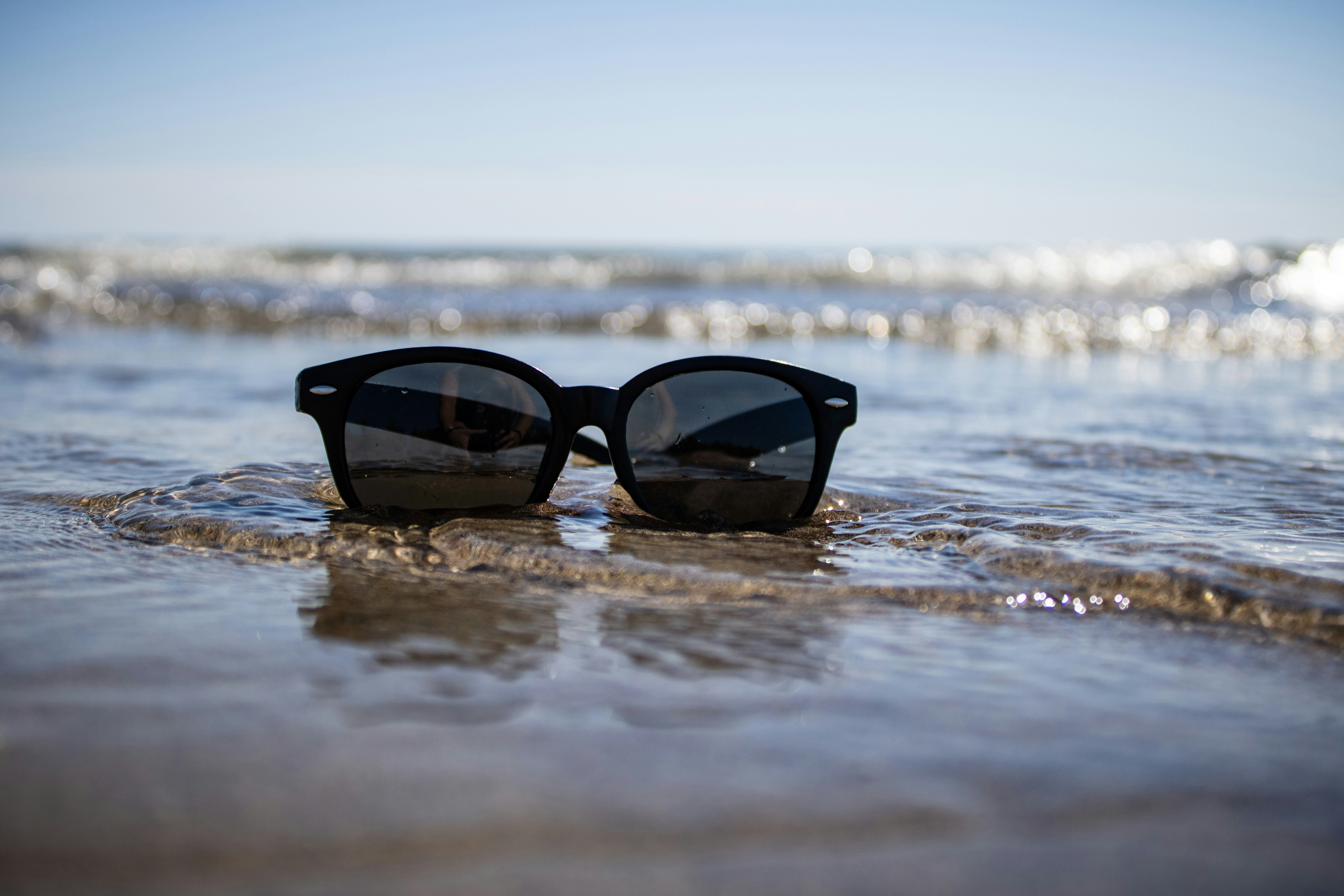 Black sunglasses resting on a sandy beach, partially submerged in gentle ocean waves. The clear blue sky reflects in the lenses.
