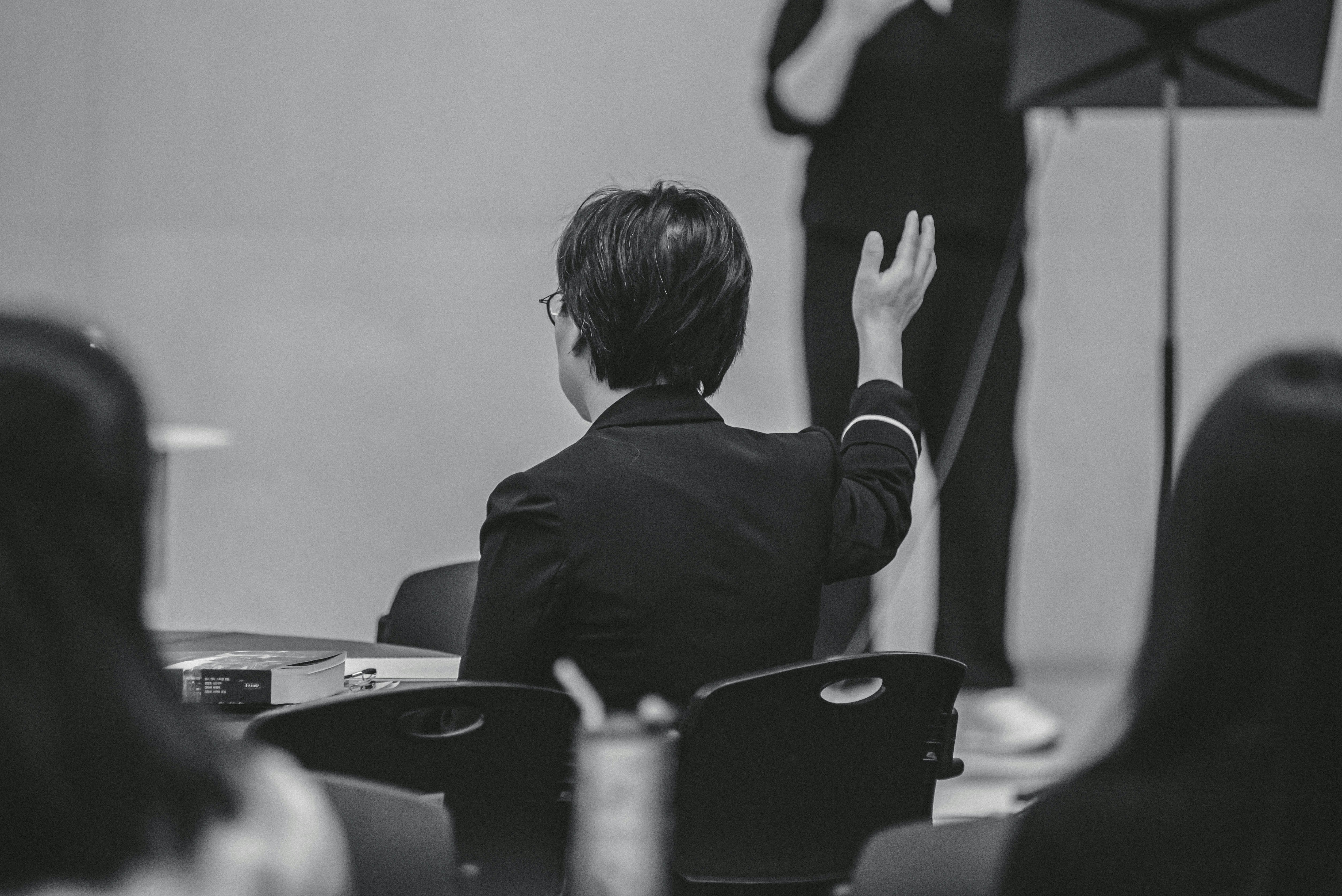 A participant in formal attire raises a hand during a discussion, embodying active engagement. The scene captures a moment of interaction in a collaborative setting.