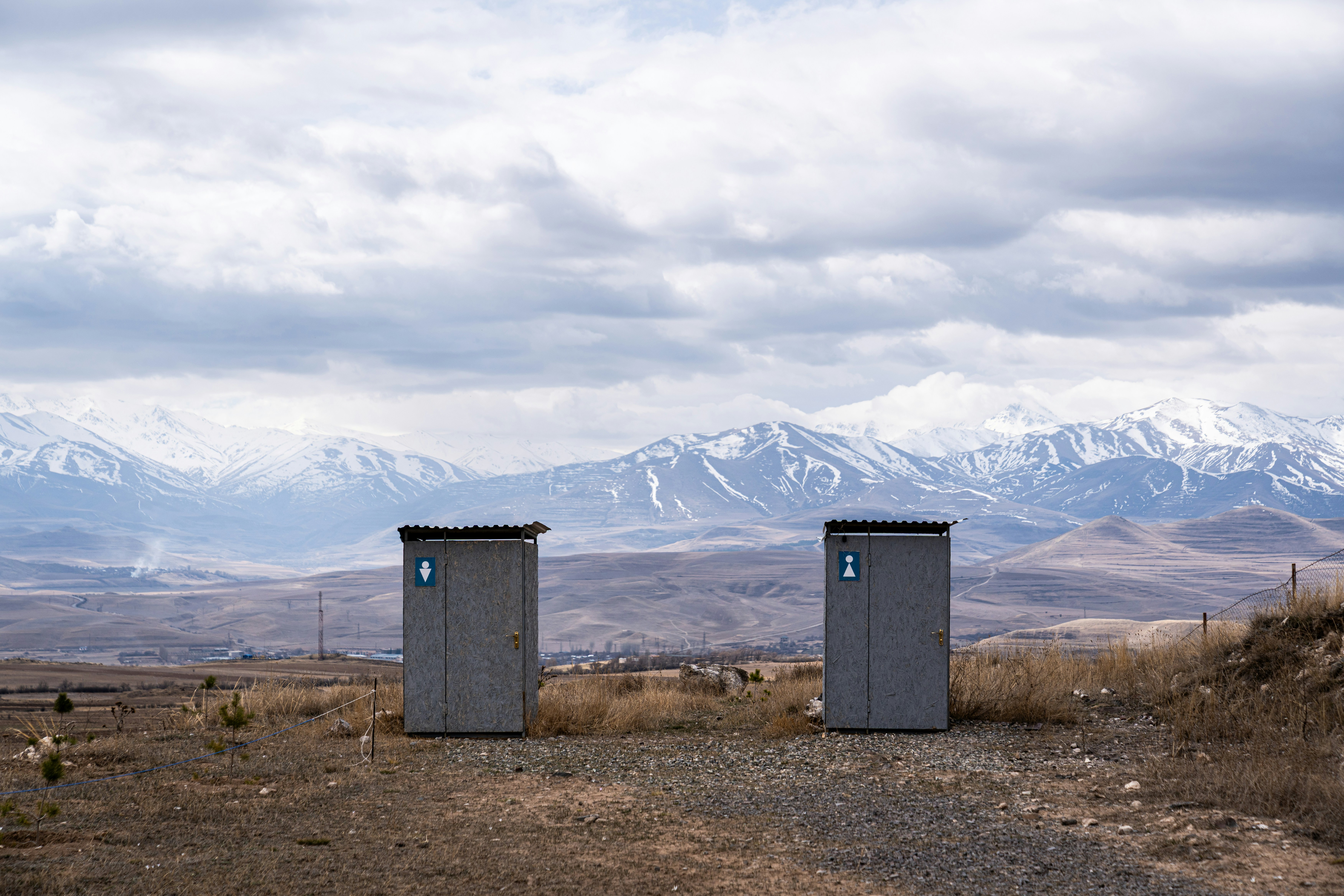 Two rustic restroom facilities stand against a backdrop of snow-capped mountains under a cloudy sky.