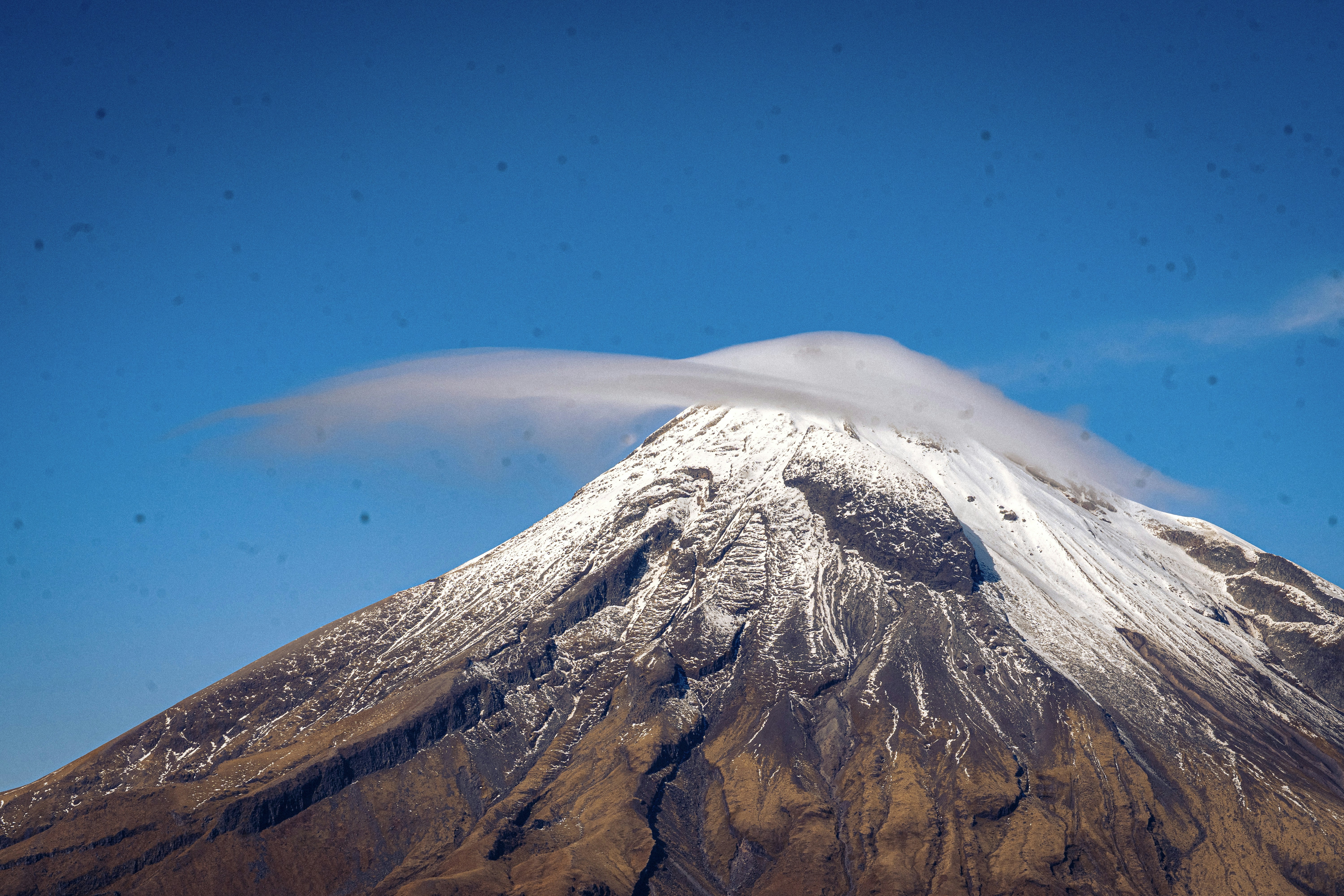 Snow-capped volcano with a unique cloud formation hovering above its peak, set against a clear blue sky.