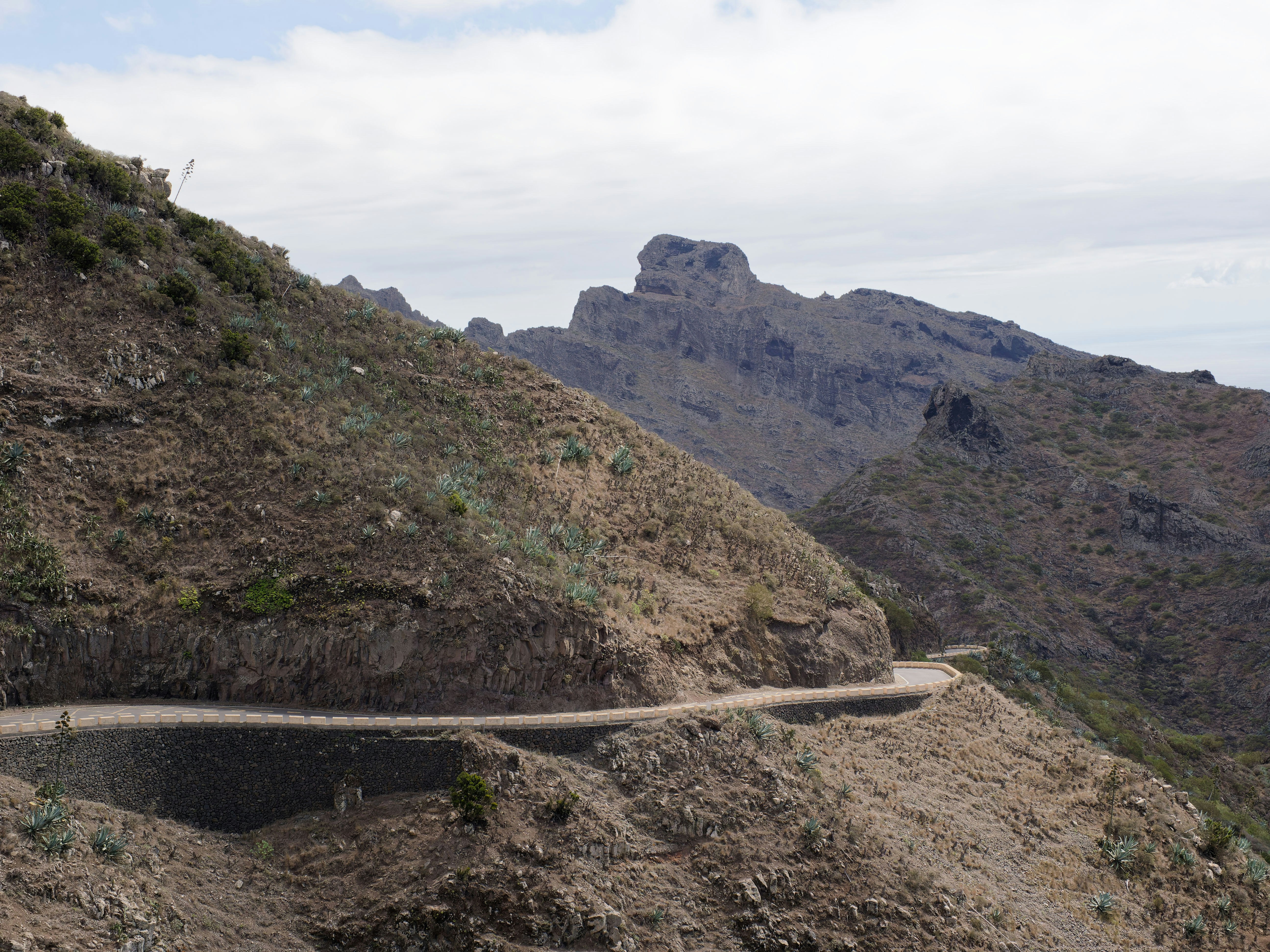 A narrow road meanders along a rocky hillside, surrounded by sparse vegetation and dramatic mountain peaks in the background.