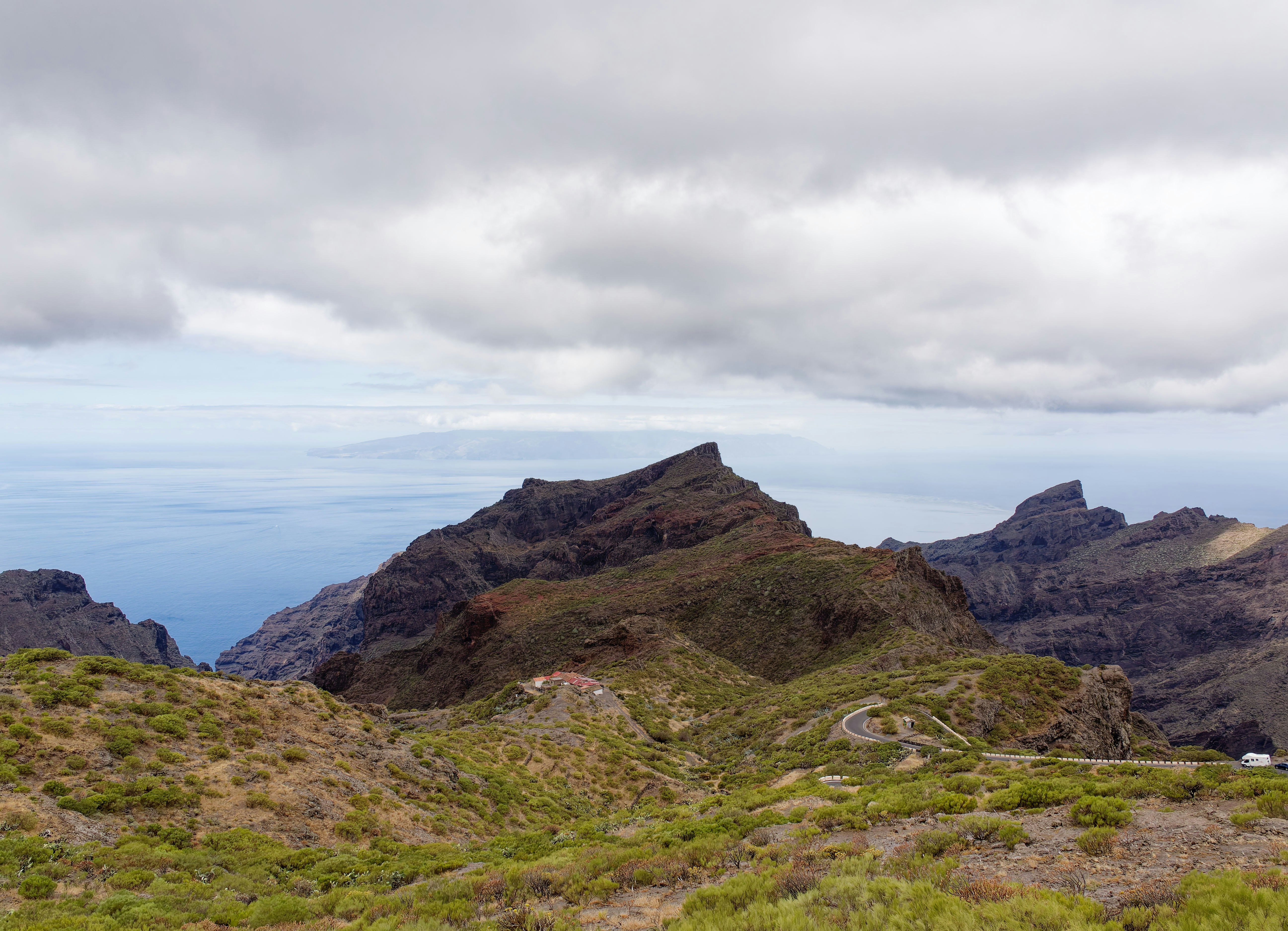 Dramatic mountain landscape overlooking a tranquil sea under a cloudy sky, showcasing winding roads and lush vegetation.