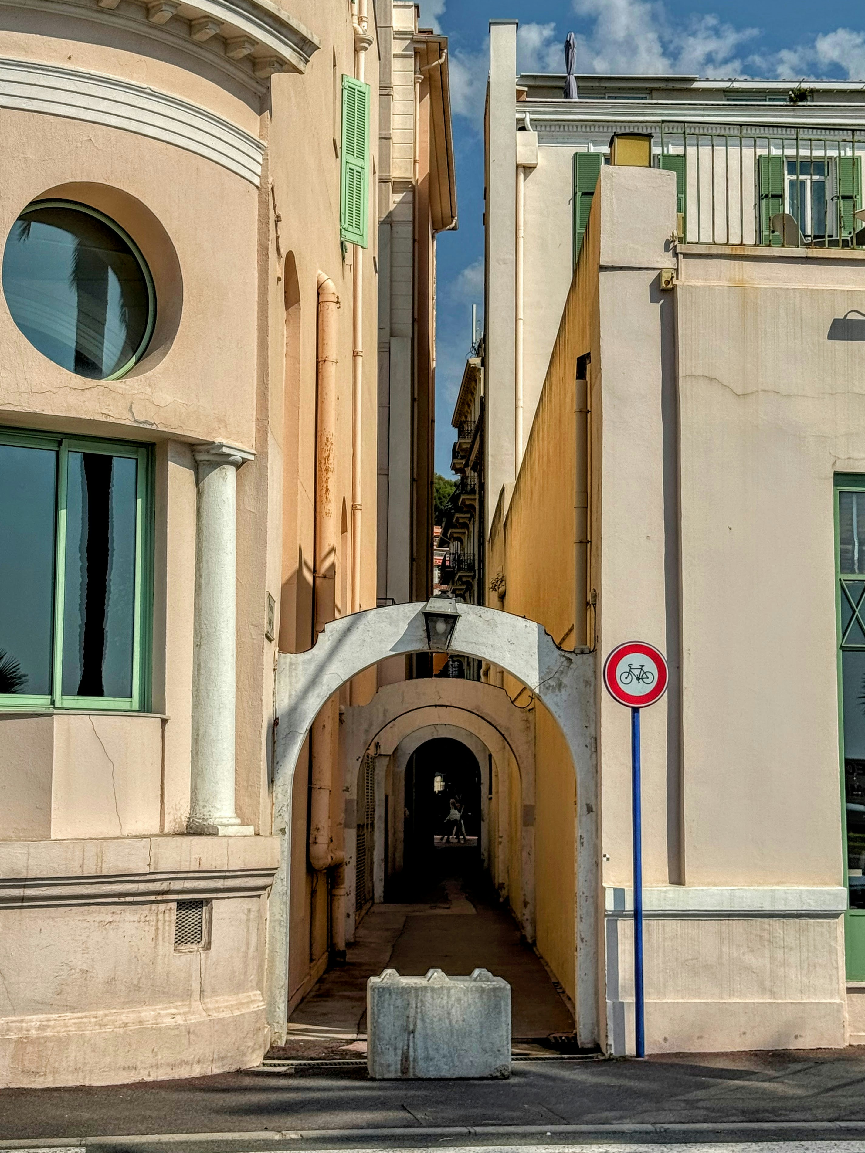 Narrow alleyway framed by pastel-colored buildings, leading to a hidden courtyard, with a bicycle sign marking the entrance.
