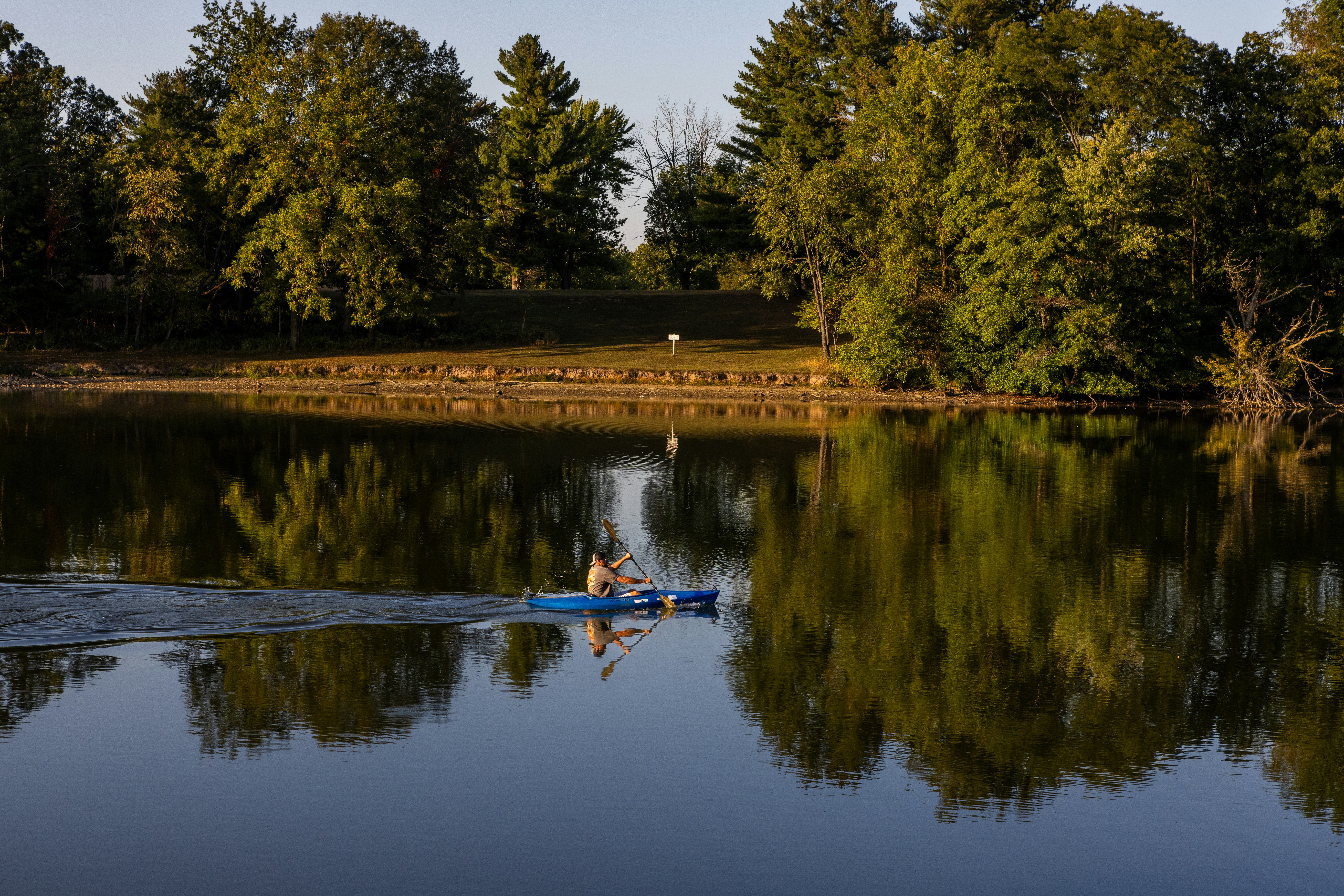 A kayaker glides across a tranquil lake, surrounded by lush greenery and mirrored reflections of the trees. The scene captures a moment of peaceful solitude.