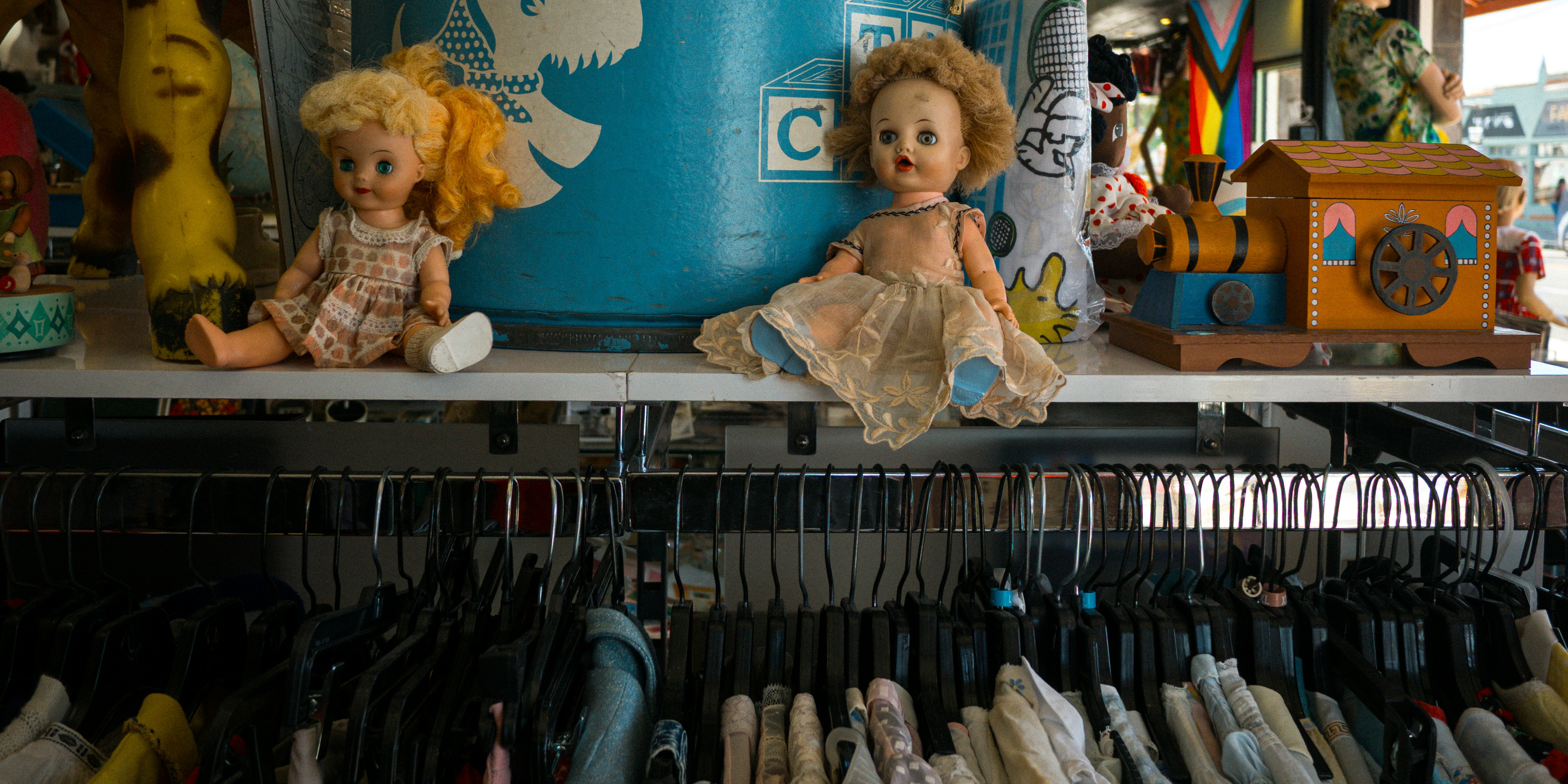 Two vintage dolls sit on a shelf above clothing racks.