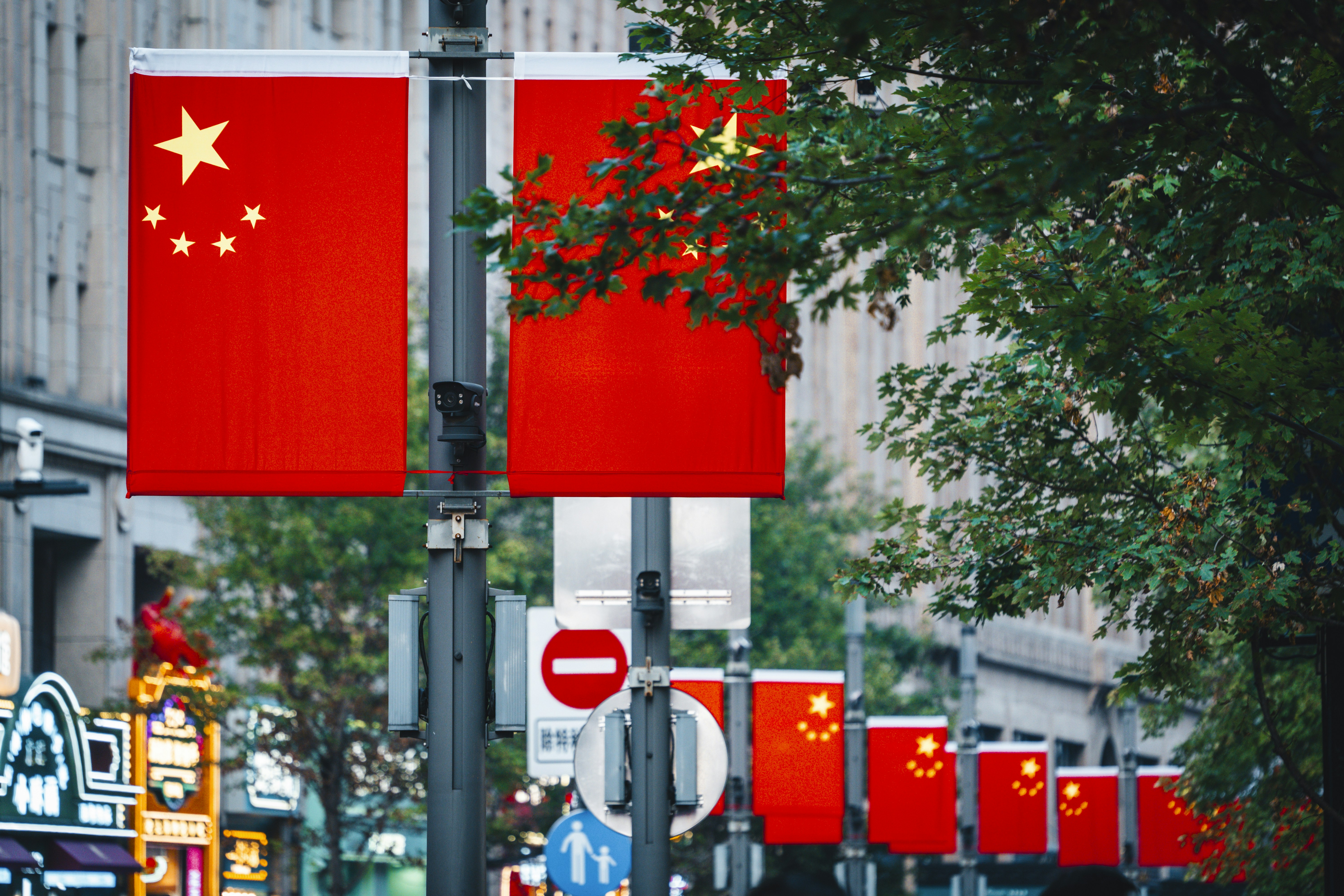 Vibrant red banners featuring the Chinese flag line a bustling street, framed by lush green foliage and urban signage.