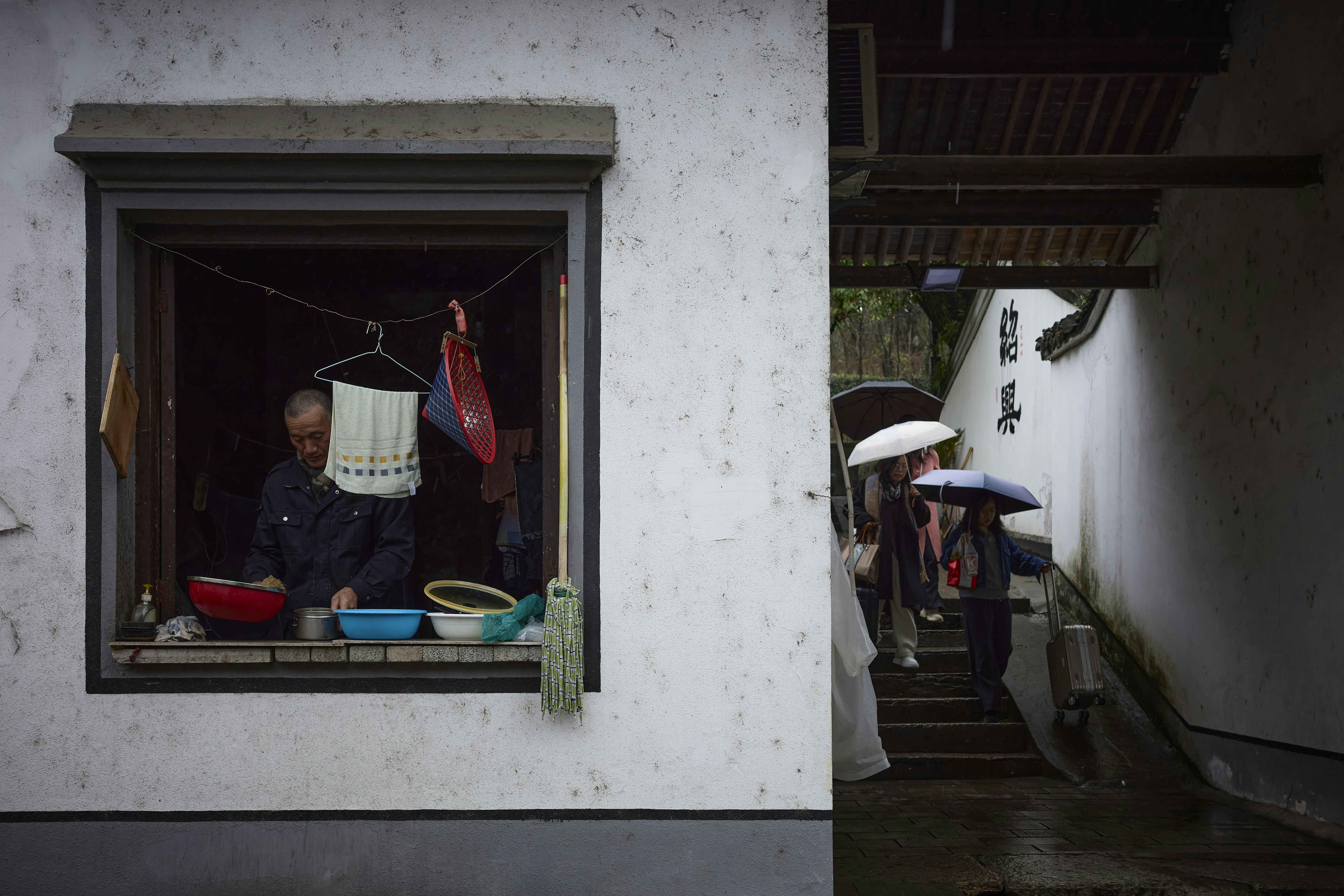 An elderly man prepares food at a window, while passersby with umbrellas navigate a rainy street in a traditional setting.