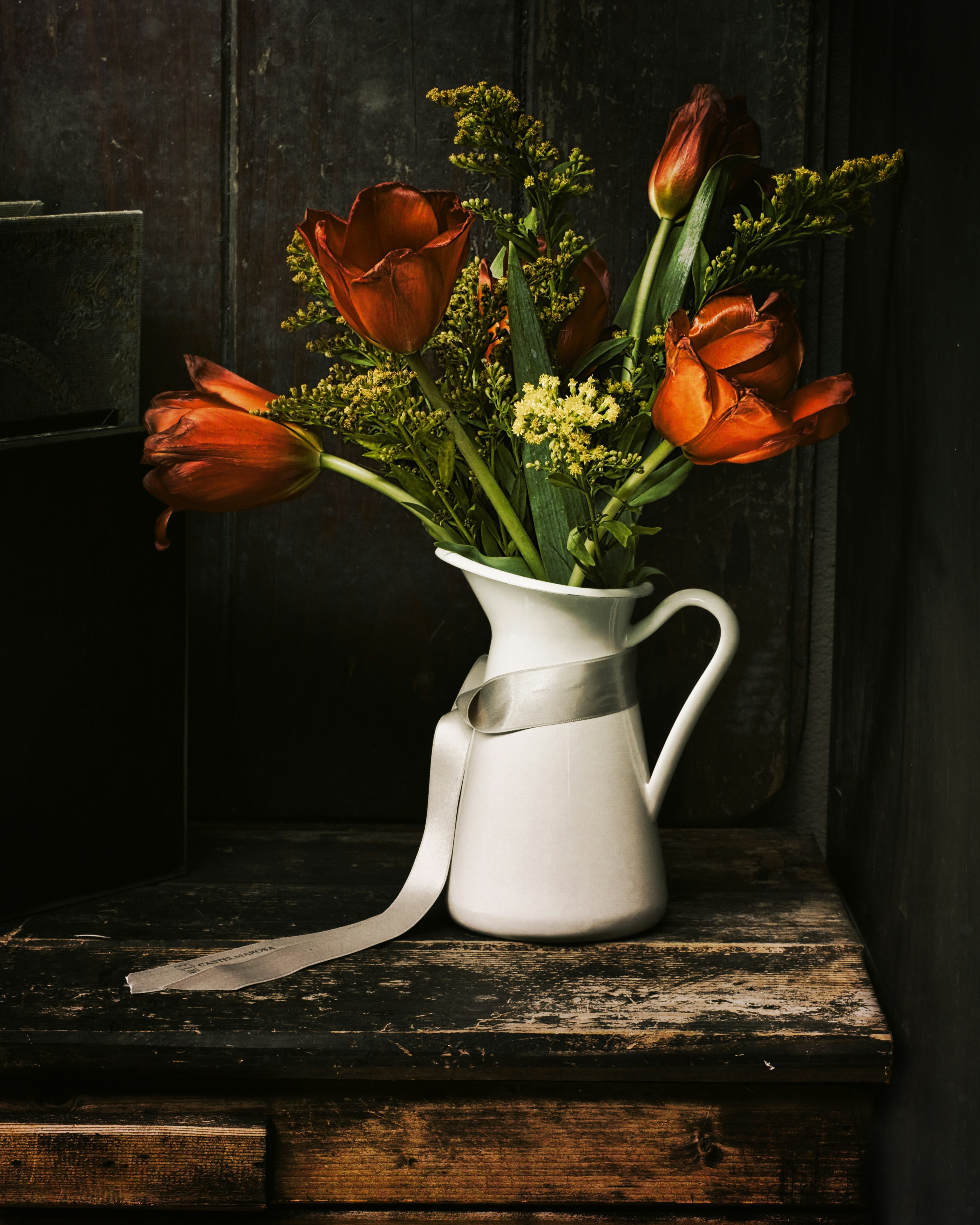 A bouquet of vibrant orange tulips and greenery arranged in a white pitcher, set against a dark wooden background.