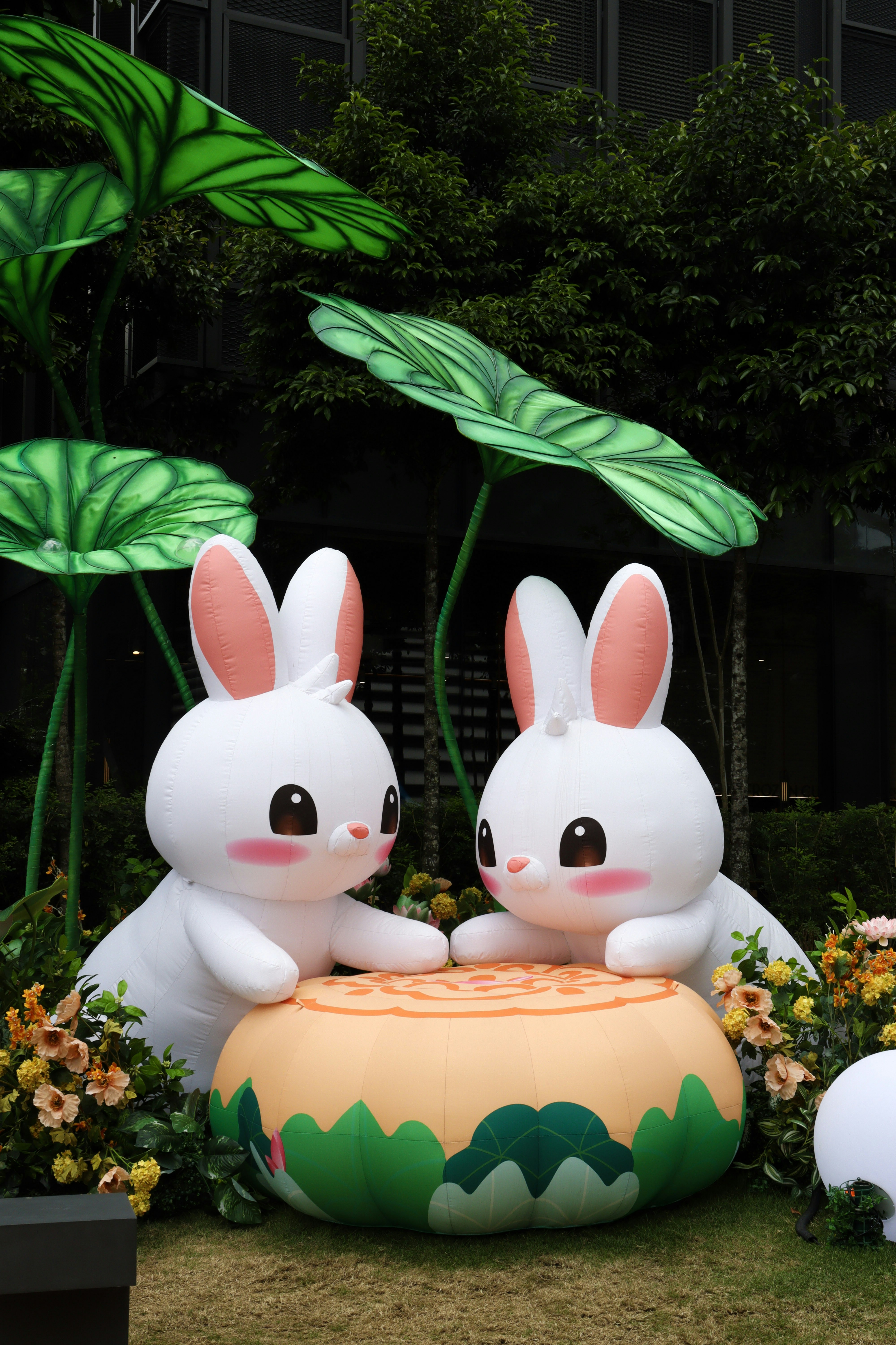 Two large, cheerful inflatable bunnies interact over a vibrant, decorative pumpkin in a lush garden setting.