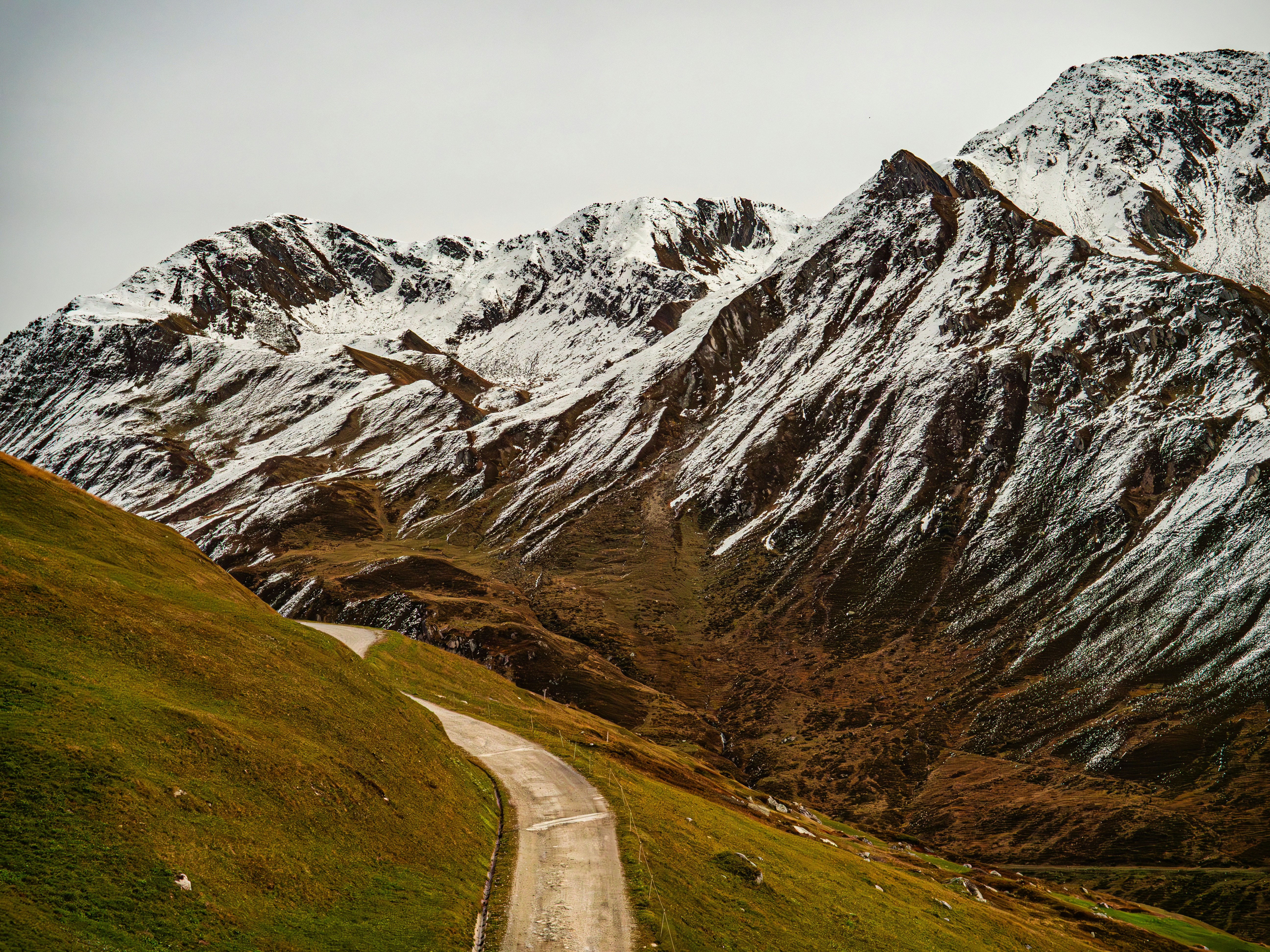 Winding dirt road leads through snow-dusted mountains, showcasing the rugged beauty of alpine terrain.