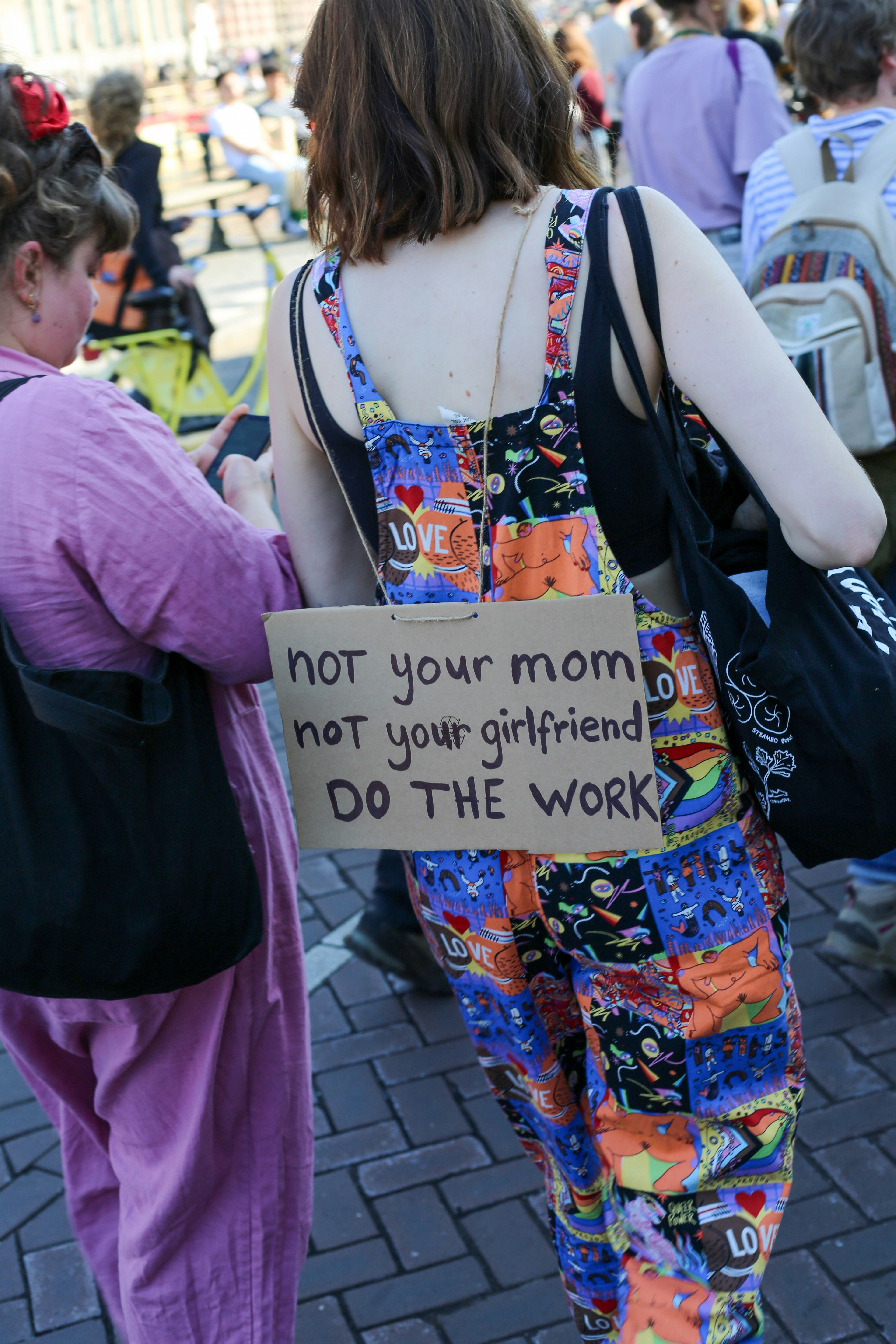 Person wearing a colorful outfit holds a sign reading 'not your mom not your girlfriend DO THE WORK' during a protest.