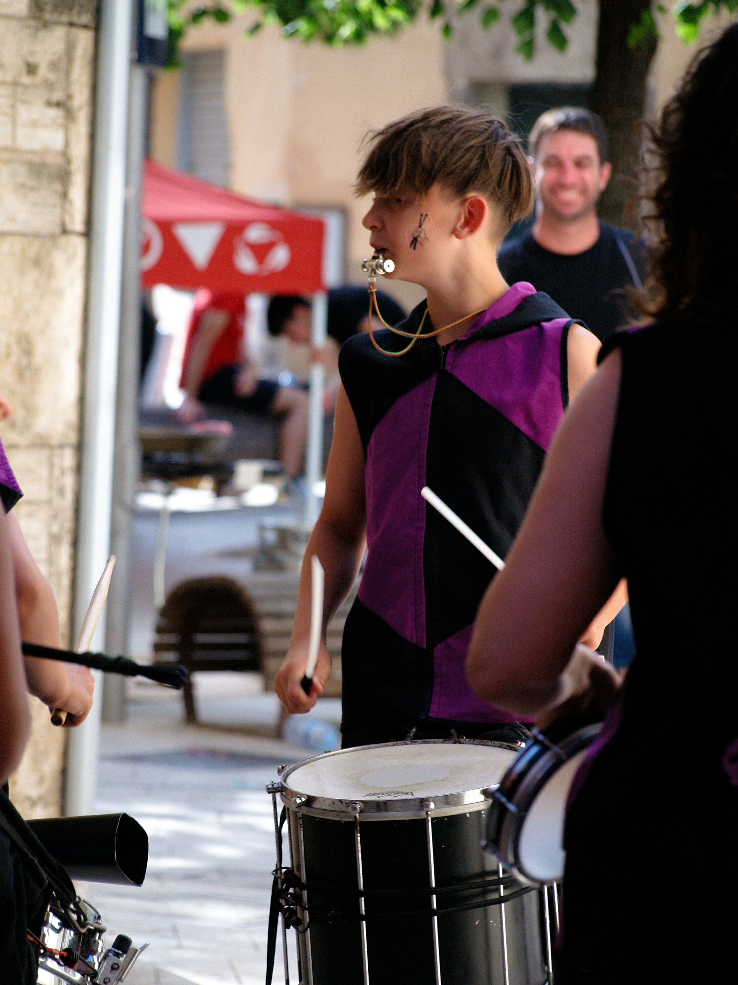 Young drummer in a vibrant outfit plays passionately in a street performance, surrounded by fellow musicians and an engaged audience. 