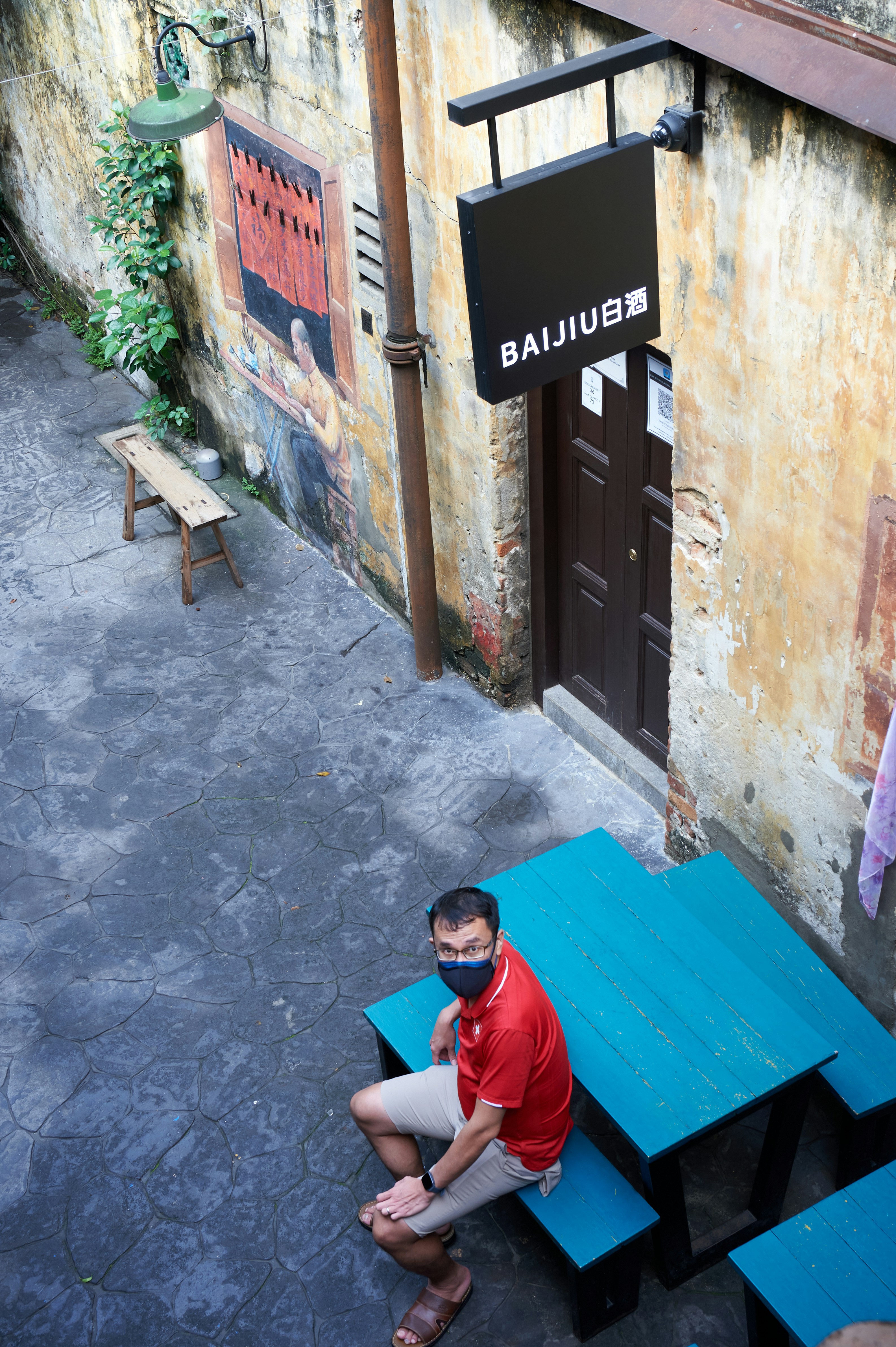 a lone guy in red t-shirt taking a rest | Man in mask sits at teal table outside building
