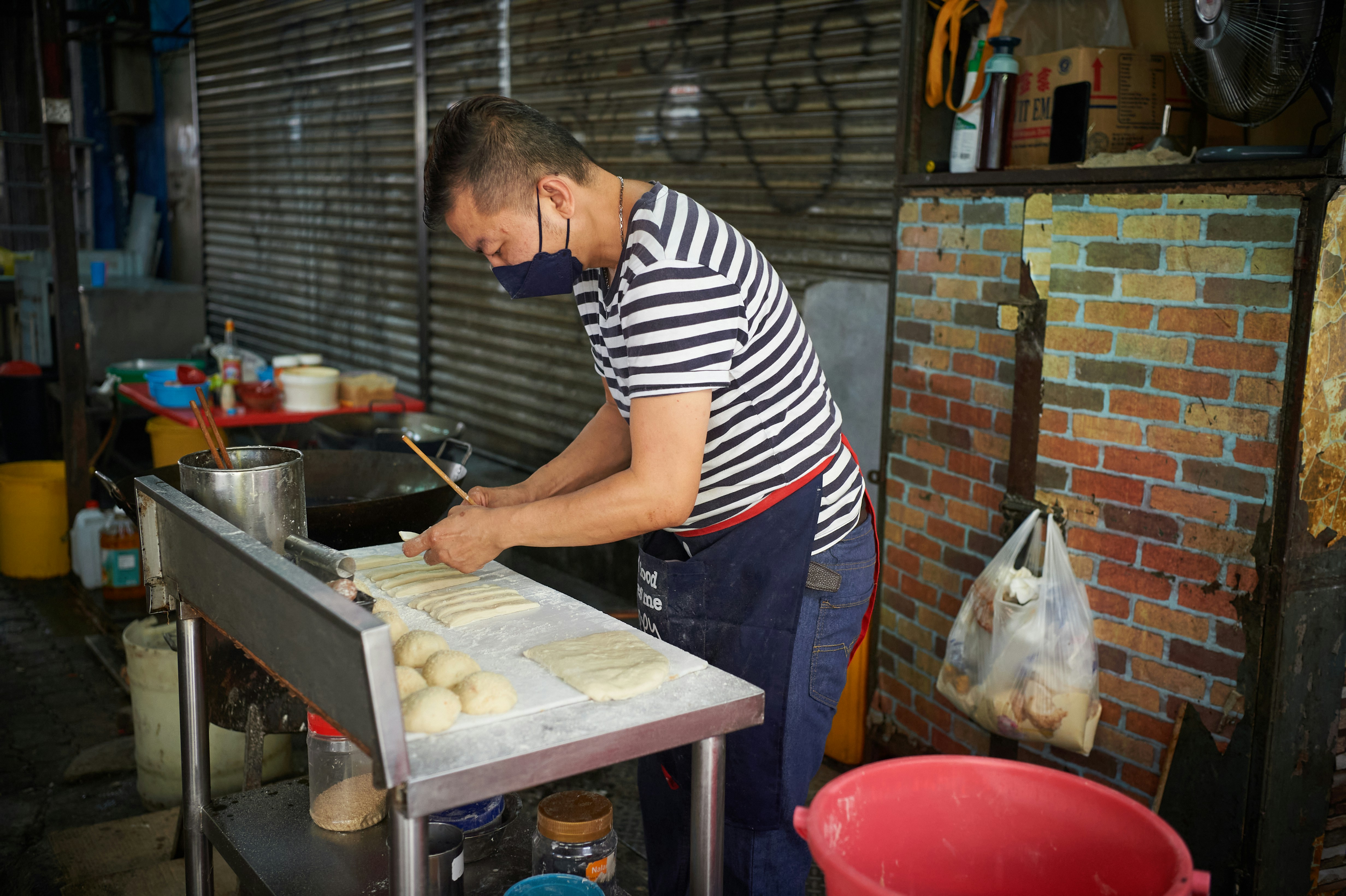 A street vendor meticulously shapes dough in a bustling kitchen, surrounded by colorful utensils and a brick wall backdrop.