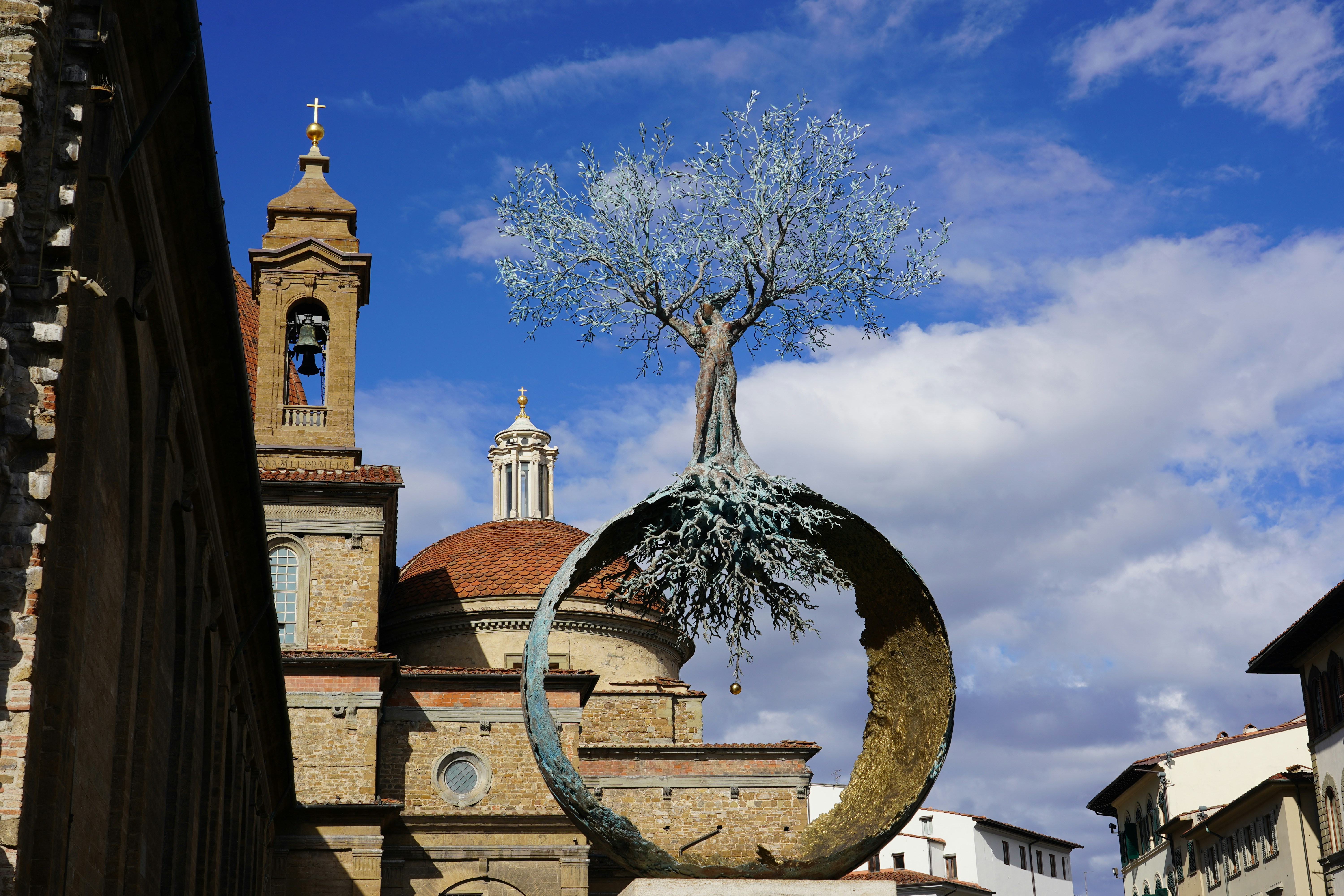A striking sculpture of a tree emerging from a circular base stands against a backdrop of historic buildings and a blue sky, symbolizing the connection between nature and urban life.