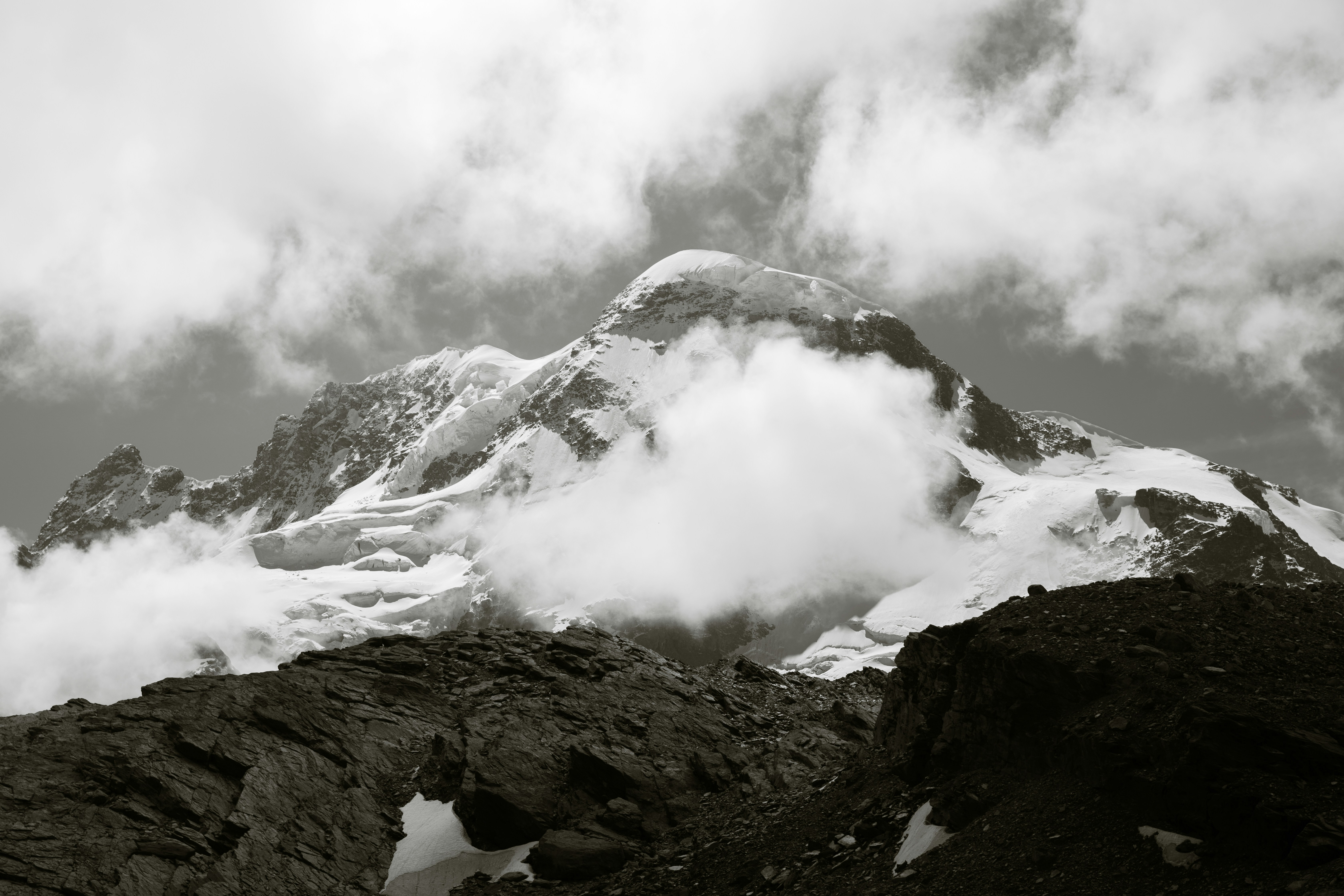Snow-capped mountain peaks partially obscured by swirling clouds, with rugged rocky foreground.