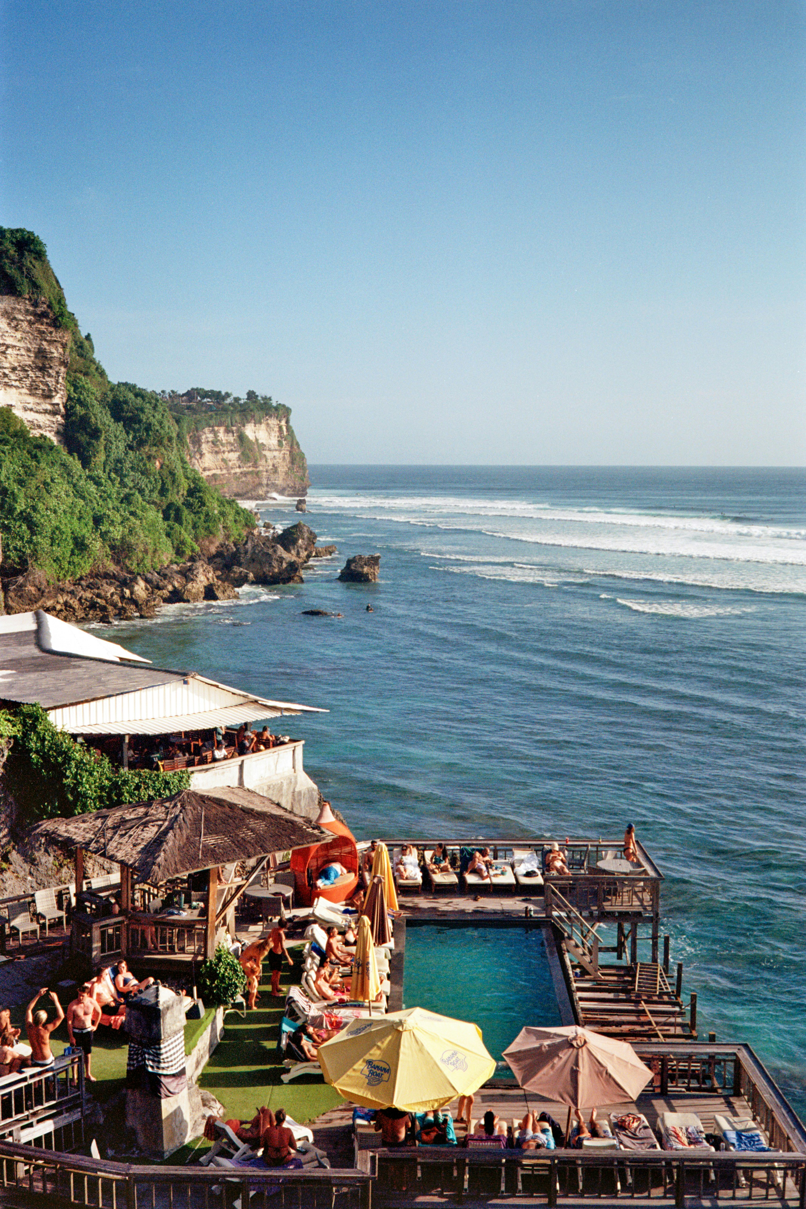Resort area with sunbathers and a pool overlooking a tranquil ocean with gentle waves. Lush greenery and cliffs frame the scene.