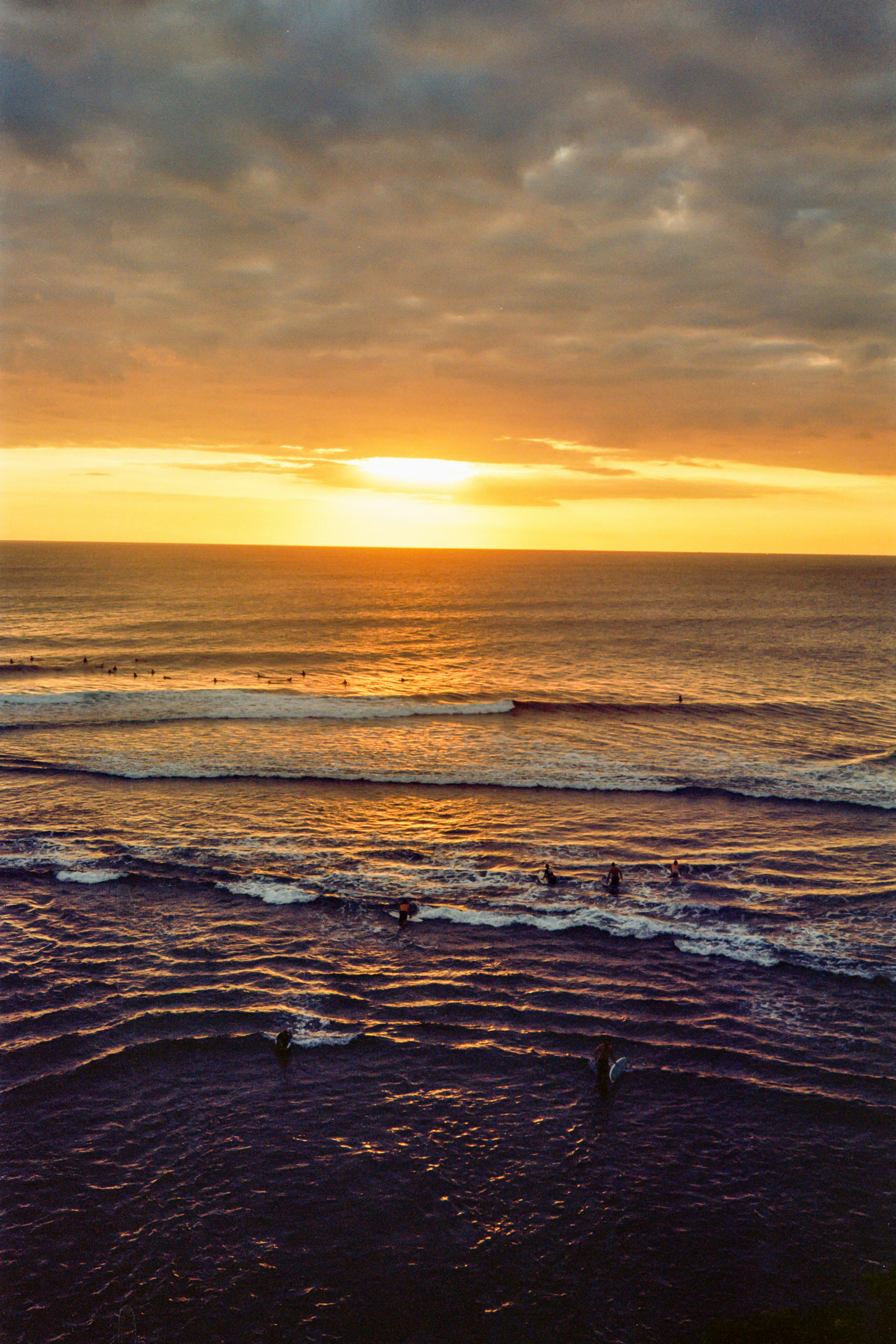 Surfers riding gentle waves as the sun sets over the horizon, casting a warm golden hue across the ocean surface.