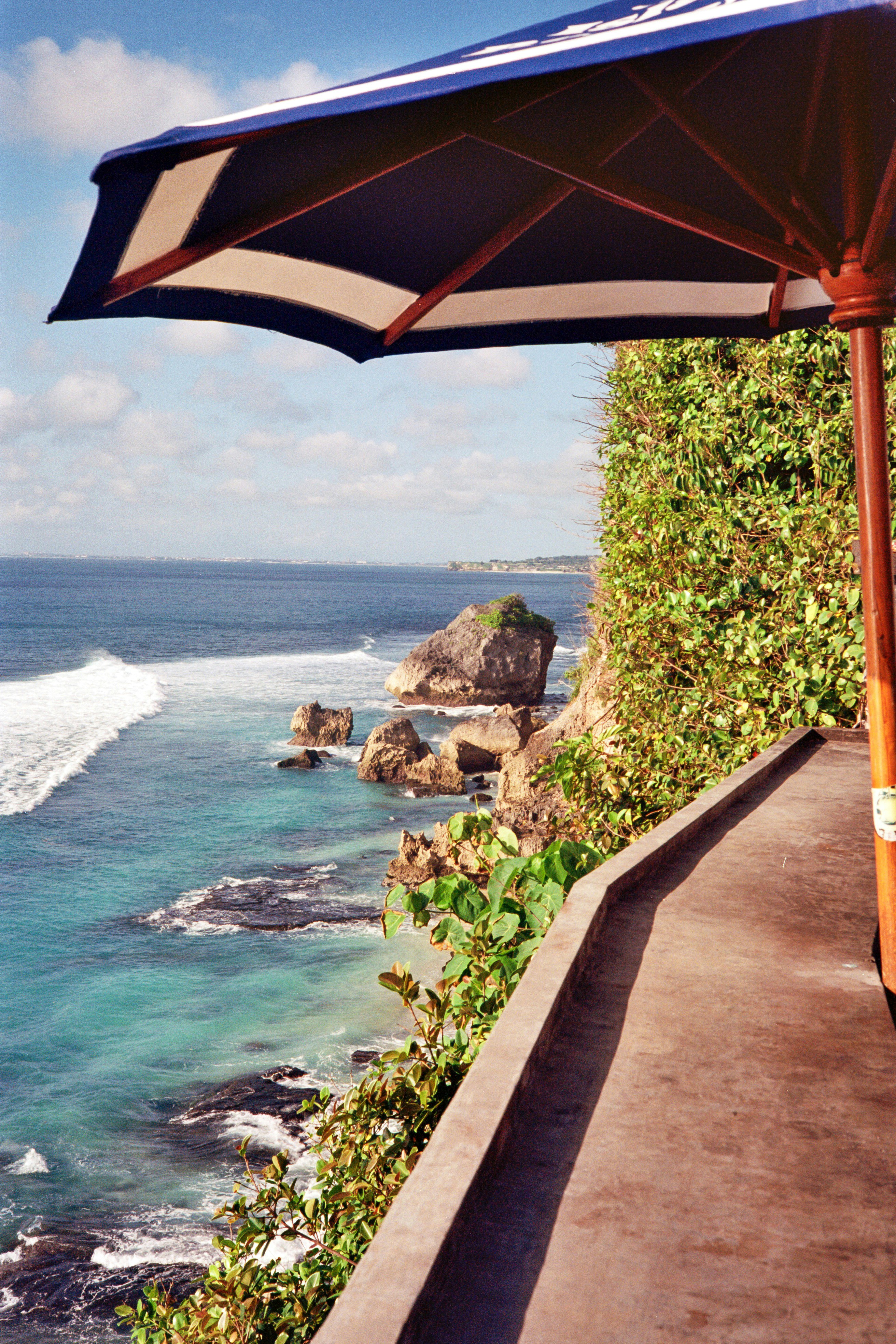 A vibrant coastal scene featuring a shaded terrace overlooking turquoise waters and rocky formations. The lush greenery adds depth to the tranquil atmosphere.