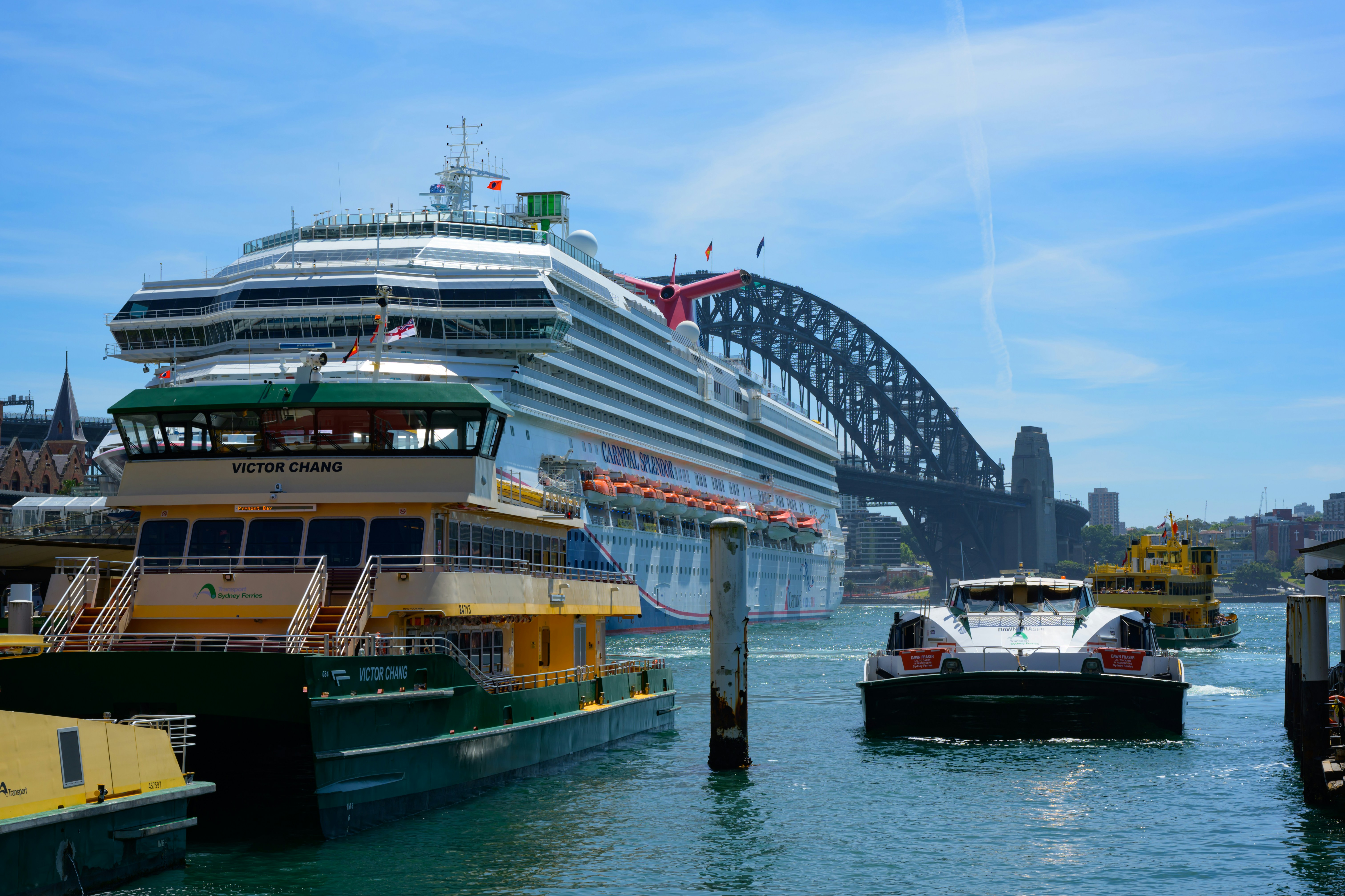 A vibrant scene showcasing a cruise ship docked alongside smaller vessels, with the iconic Sydney Harbour Bridge in the background. The clear blue sky enhances the lively atmosphere.