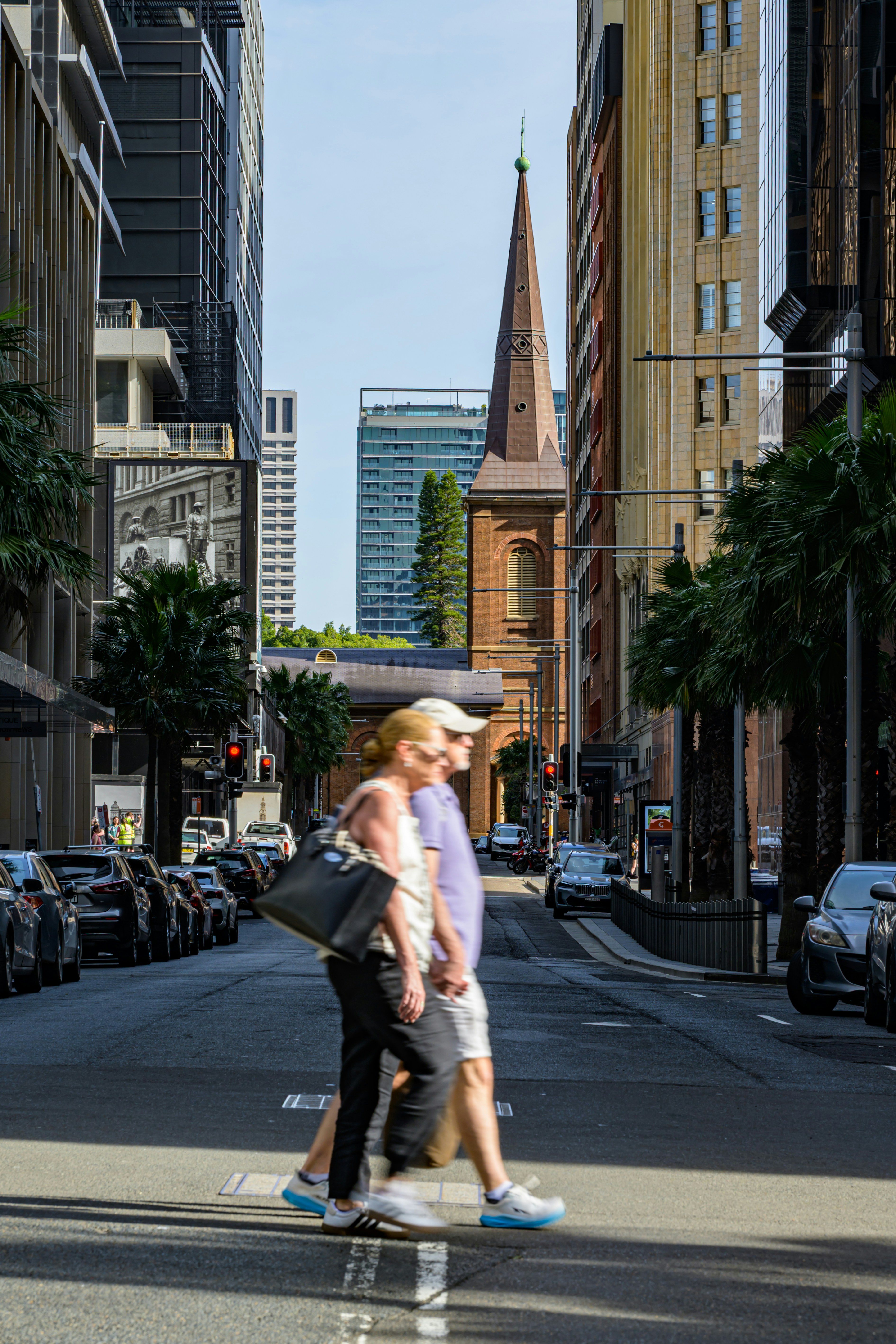 Pedestrians crossing a city street with a historic church steeple visible in the background among modern skyscrapers. Palm trees line the road, adding a touch of greenery.