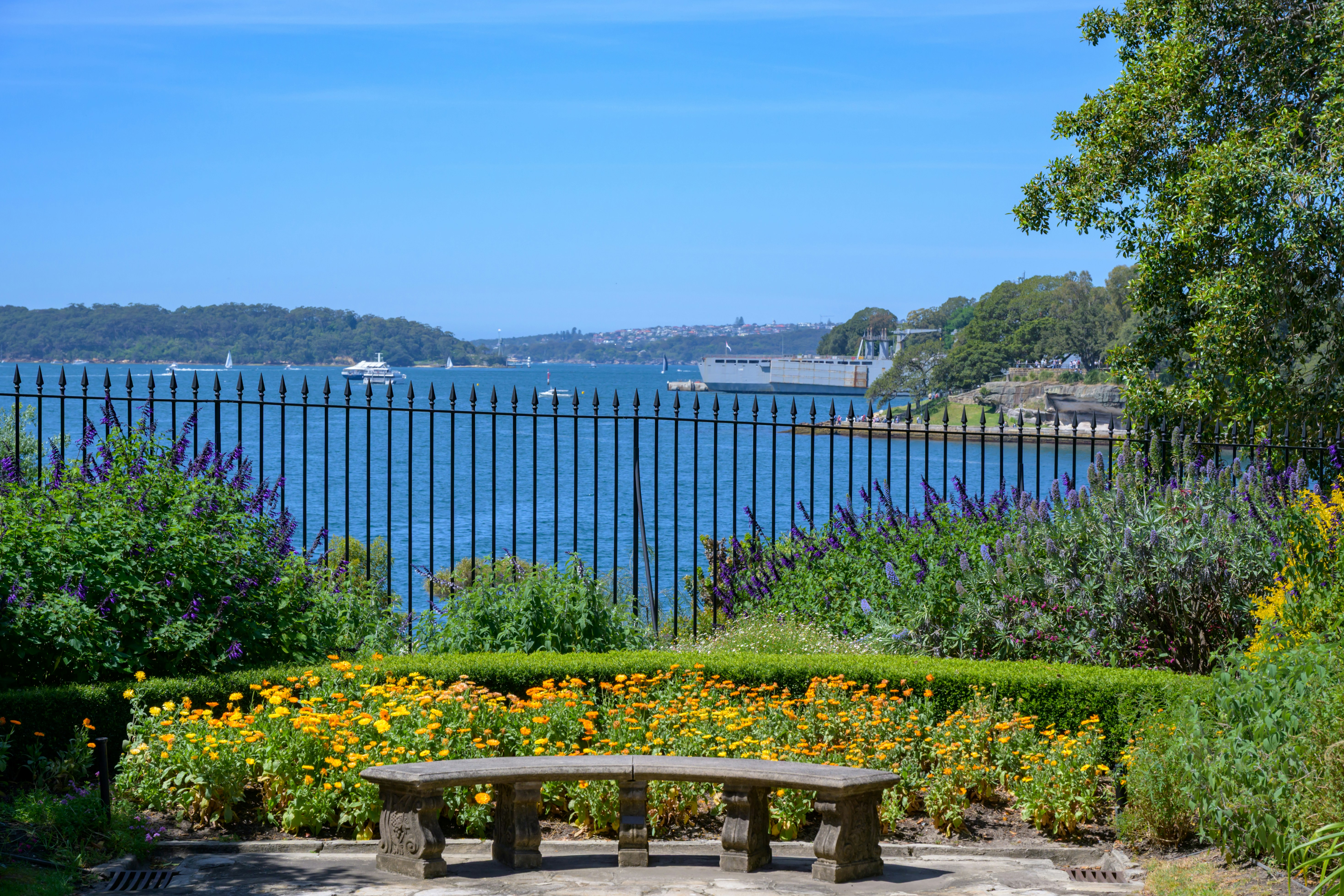 Vibrant garden bench framed by colorful flowers overlooking a tranquil bay with boats in the distance.