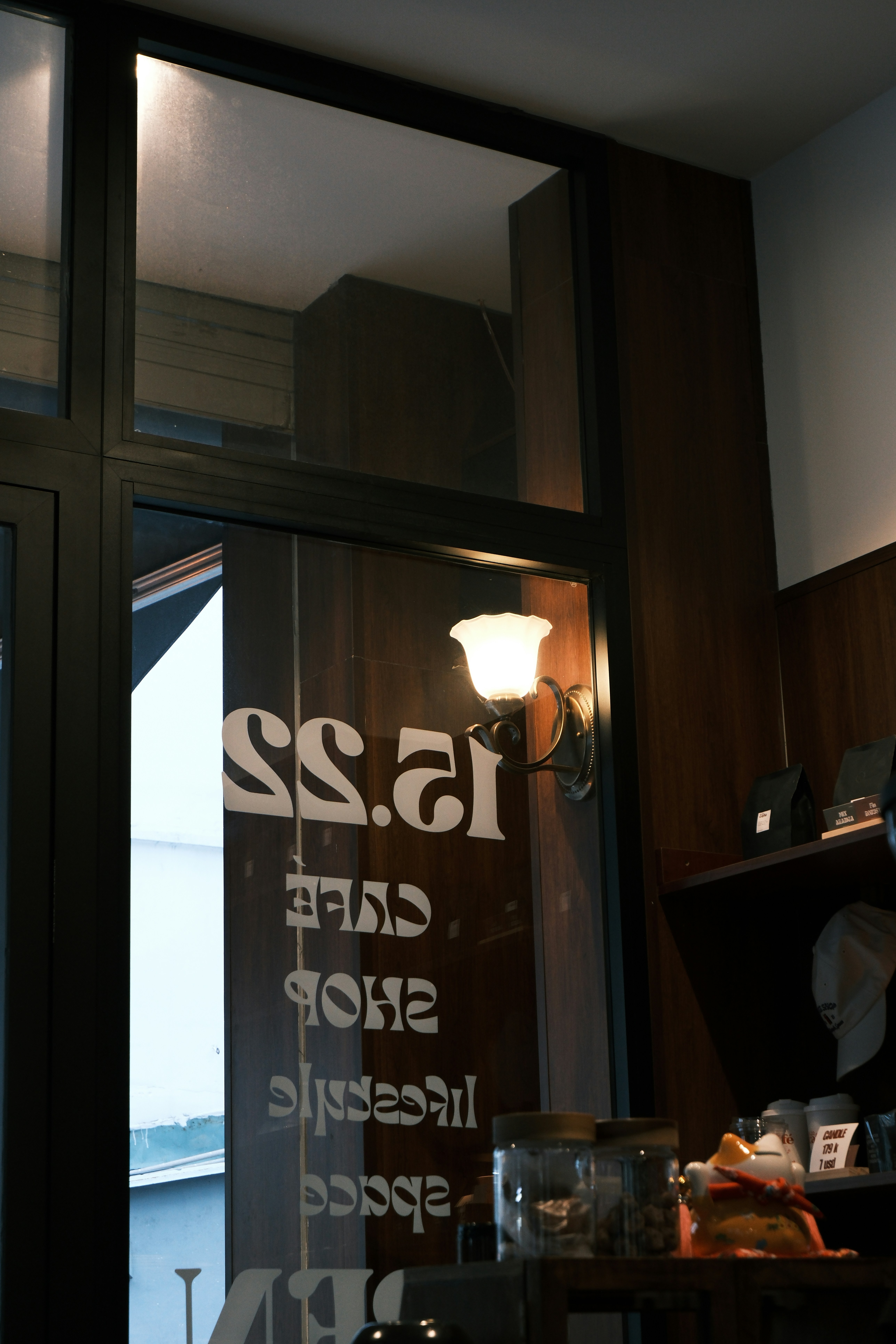 A warm light fixture illuminates a café's window, showcasing the shop's name and a glimpse of the interior decor. The scene captures a blend of inviting ambiance and urban charm.