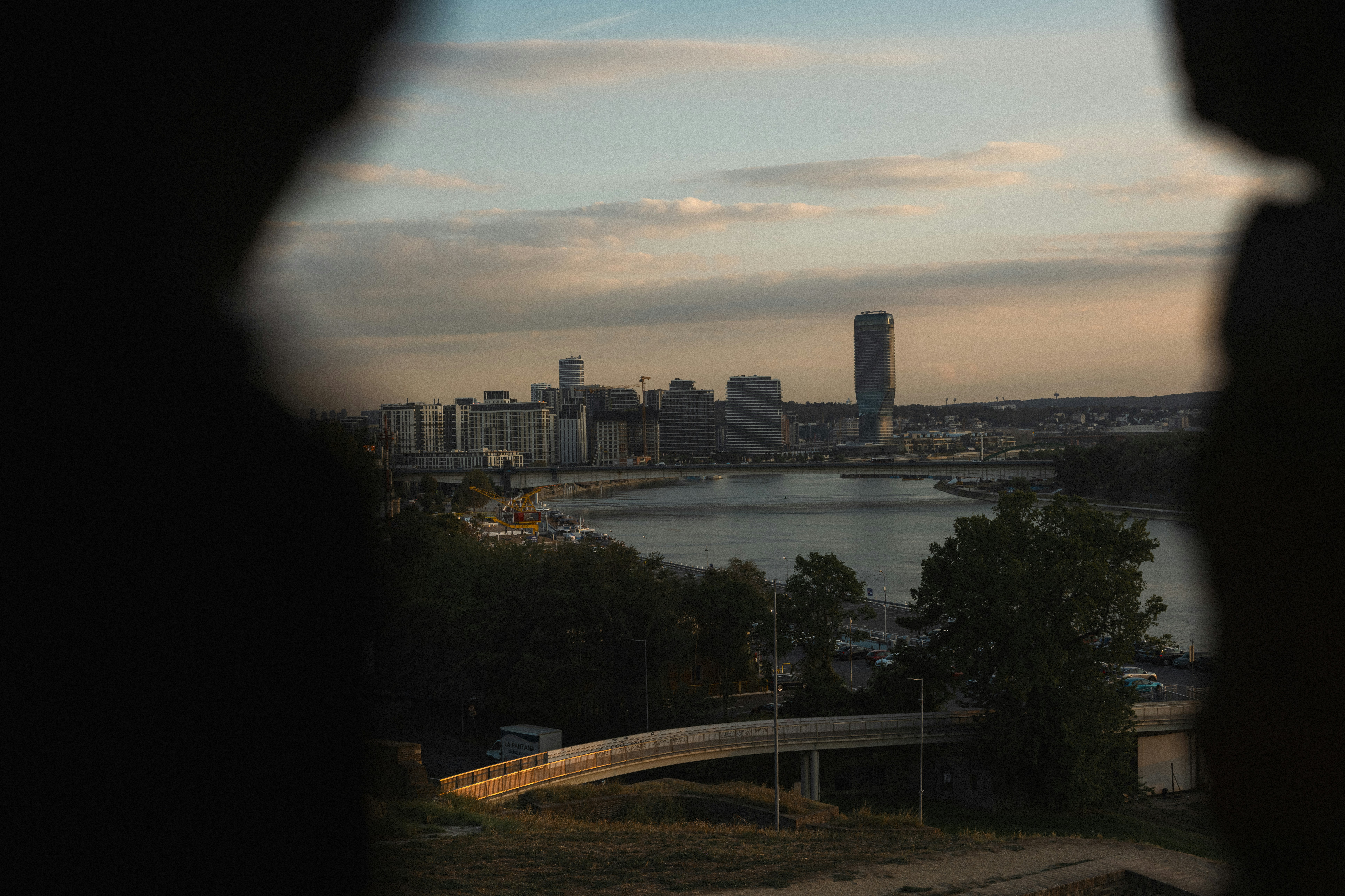 City skyline peeks through a natural frame, showcasing a tranquil river and modern architecture at dusk.