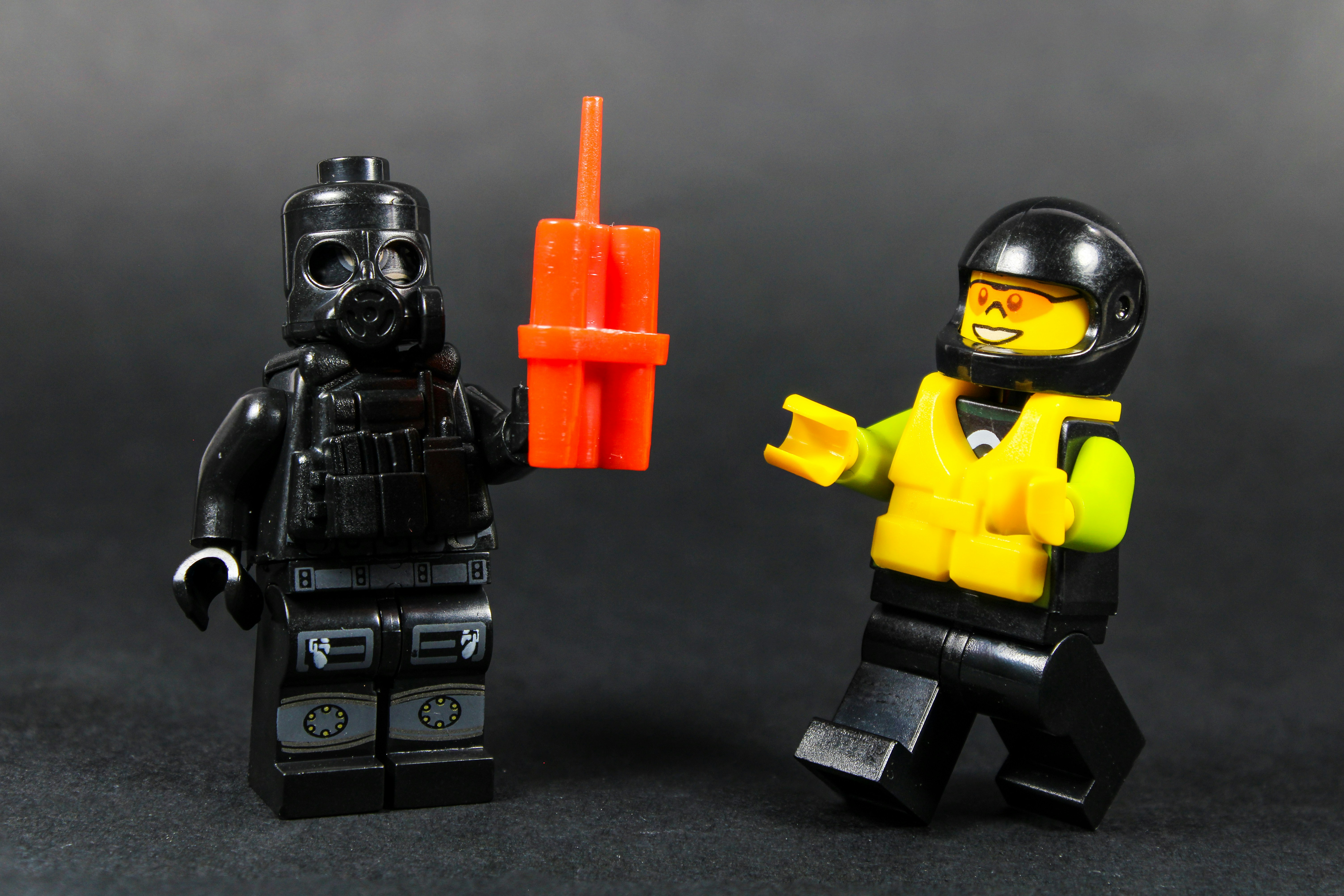 Two LEGO figures engage in a playful exchange, one holding an orange object while the other gestures enthusiastically. The scene is set against a dark background.