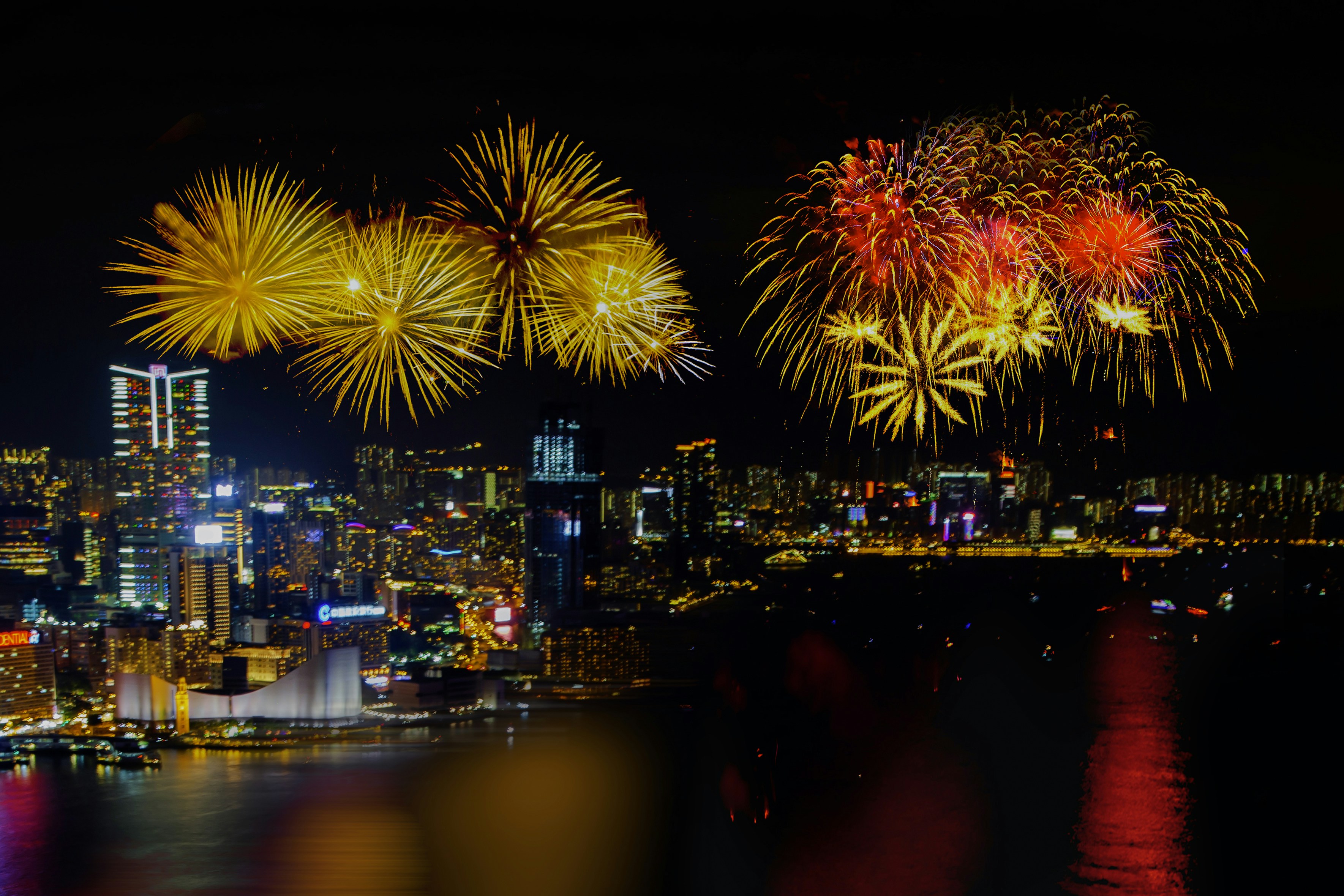 Vibrant fireworks burst above a dazzling city skyline, reflecting in the water below. The scene captures the energy and celebration of a festive night.