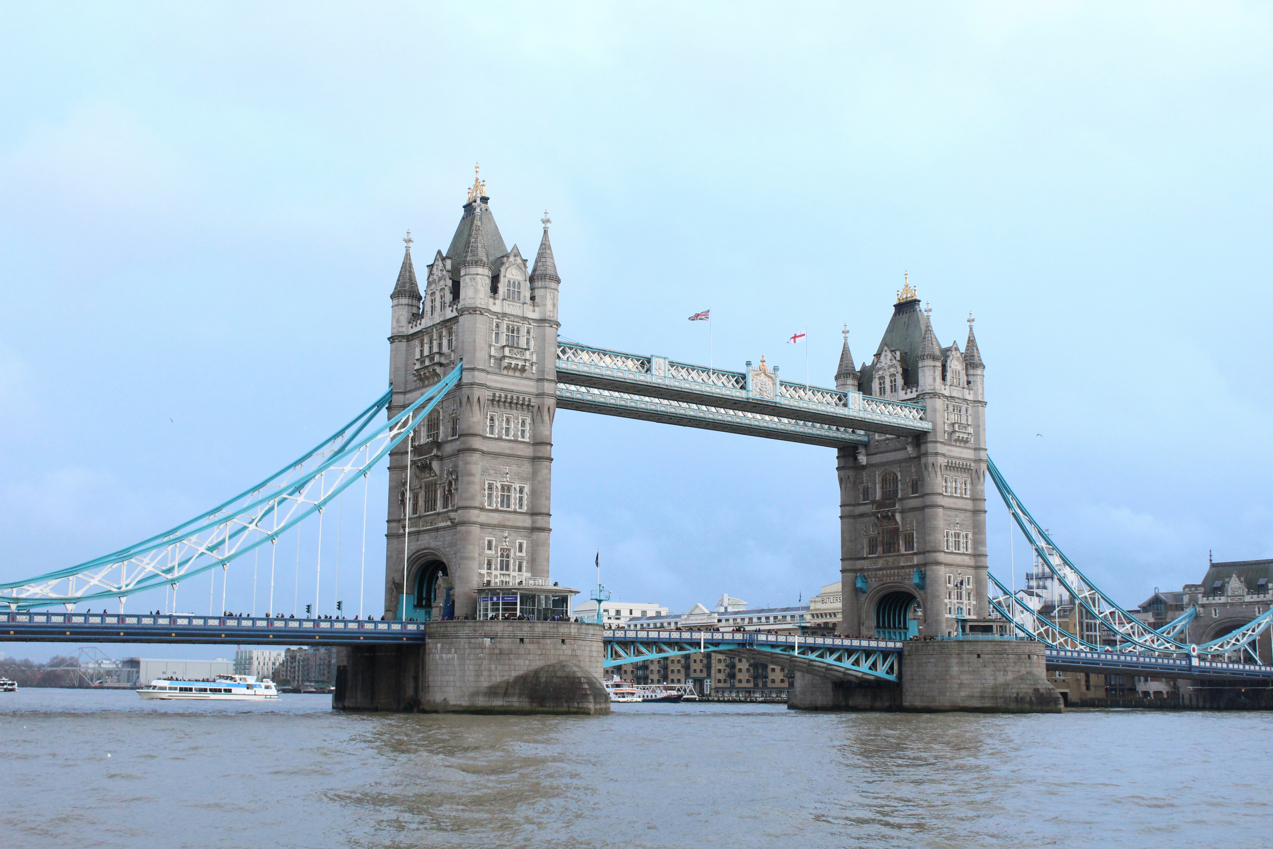 Tower Bridge spans the River Thames, showcasing its distinctive Gothic architecture and vibrant blue accents. The scene captures the bridge's grandeur against a cloudy sky.